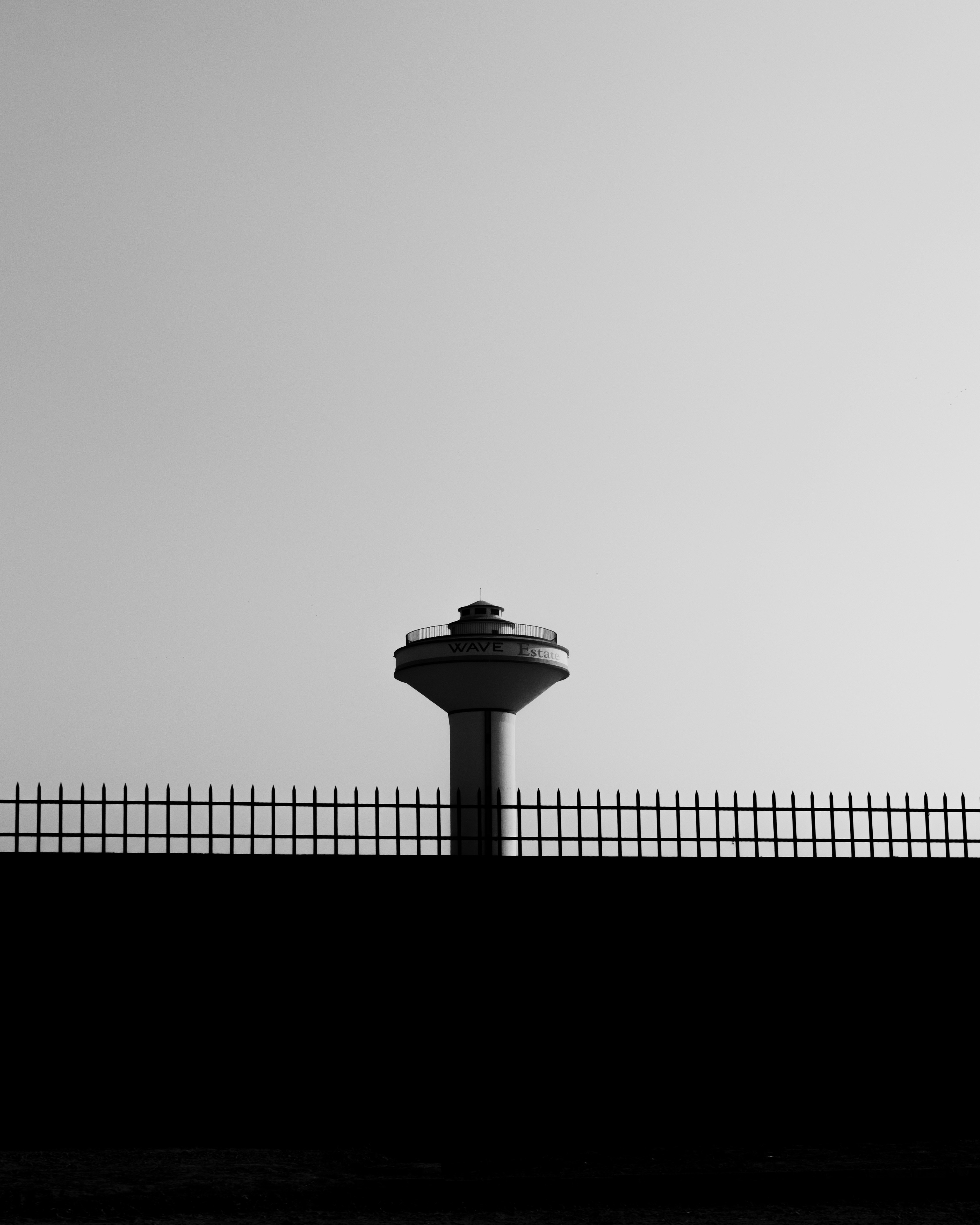Silhouette of a water tower against a gradient sky, framed by a stark fence. The minimalist composition emphasizes the tower's structure.