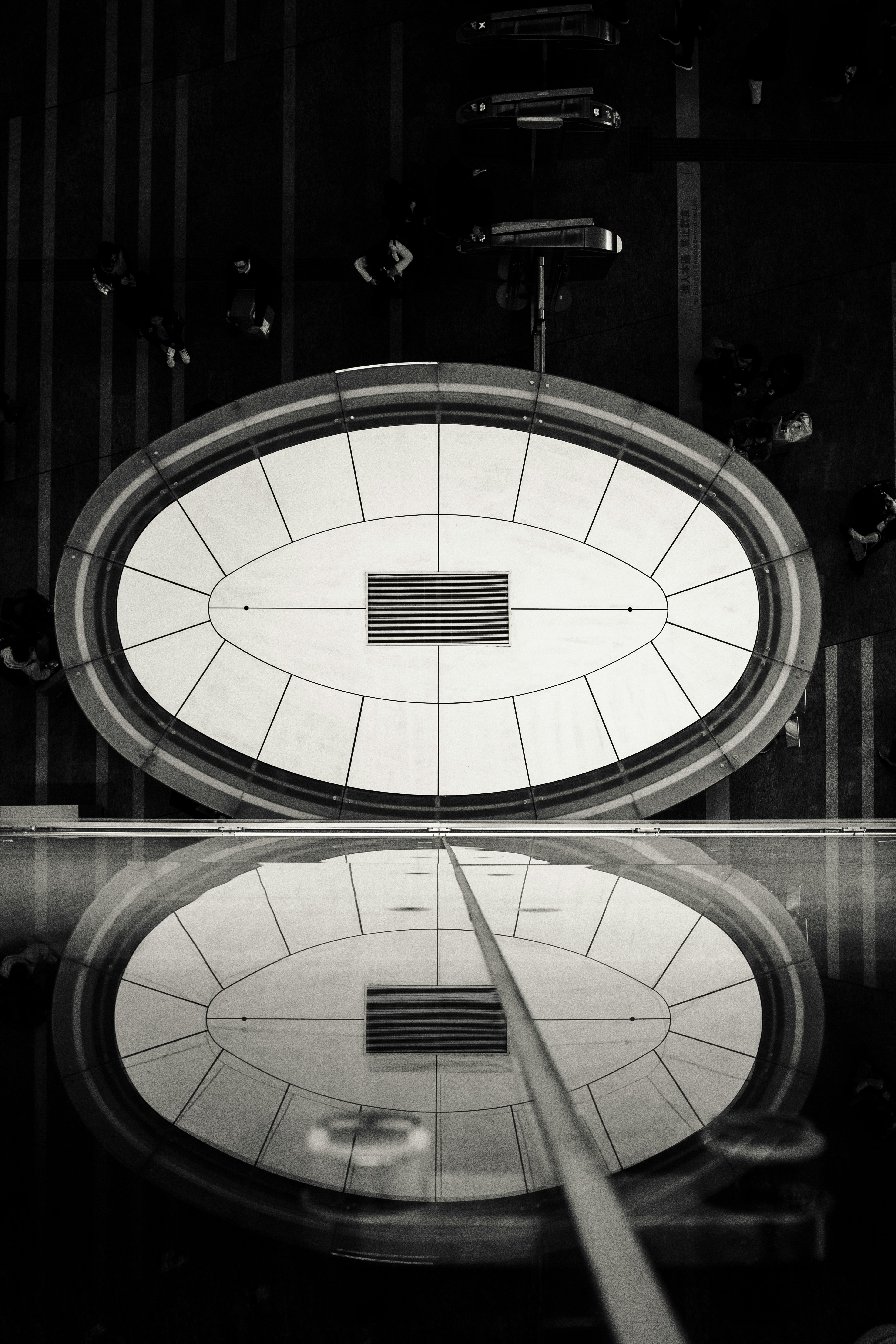 Aerial view of an elliptical architectural feature reflecting on polished surface, surrounded by bustling figures below.