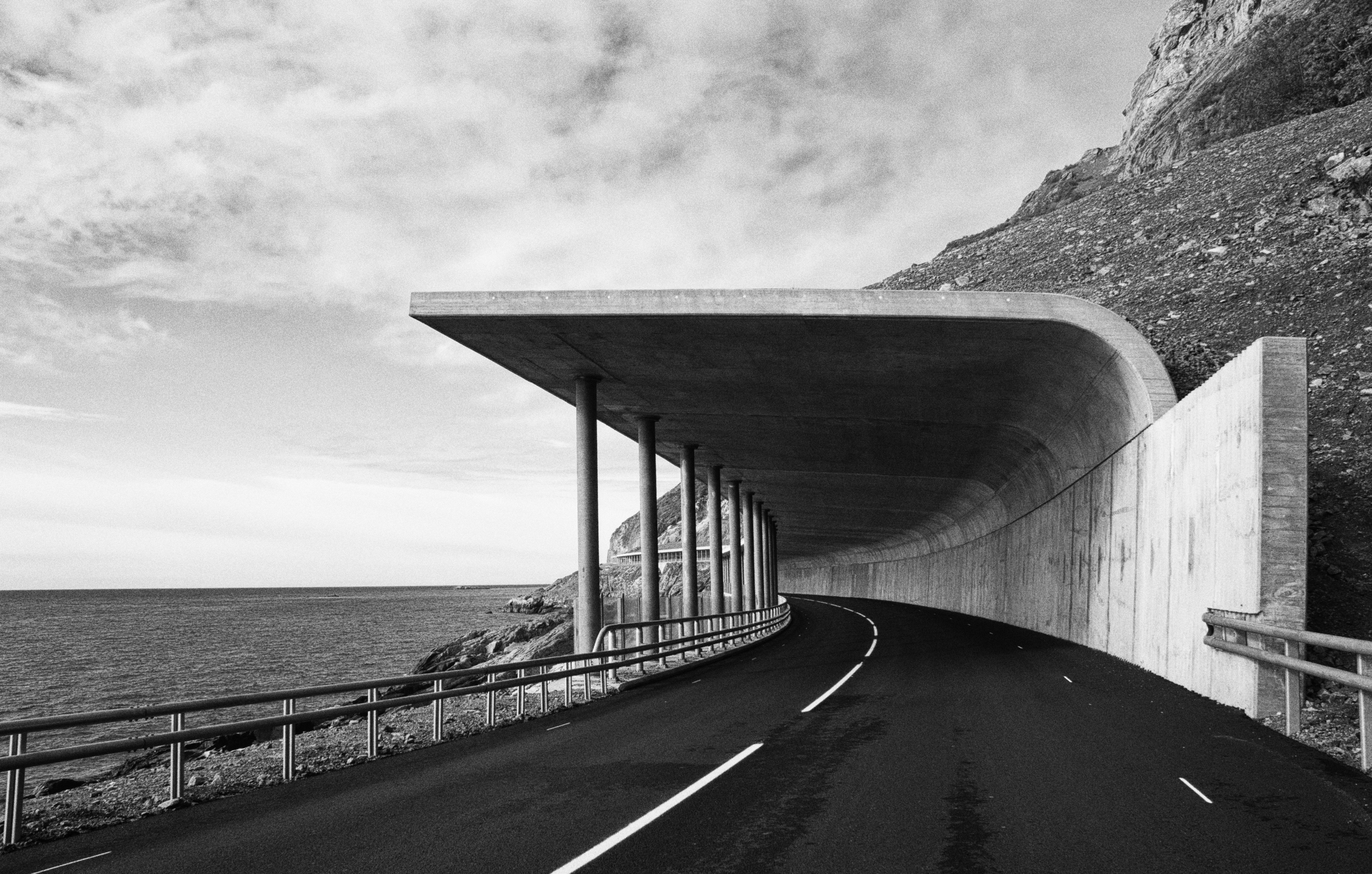 Monochrome coastal road bends beneath a concrete canopy along a cliff, curving toward the horizon. This stark architectural composition relies on bold lines and negative space.