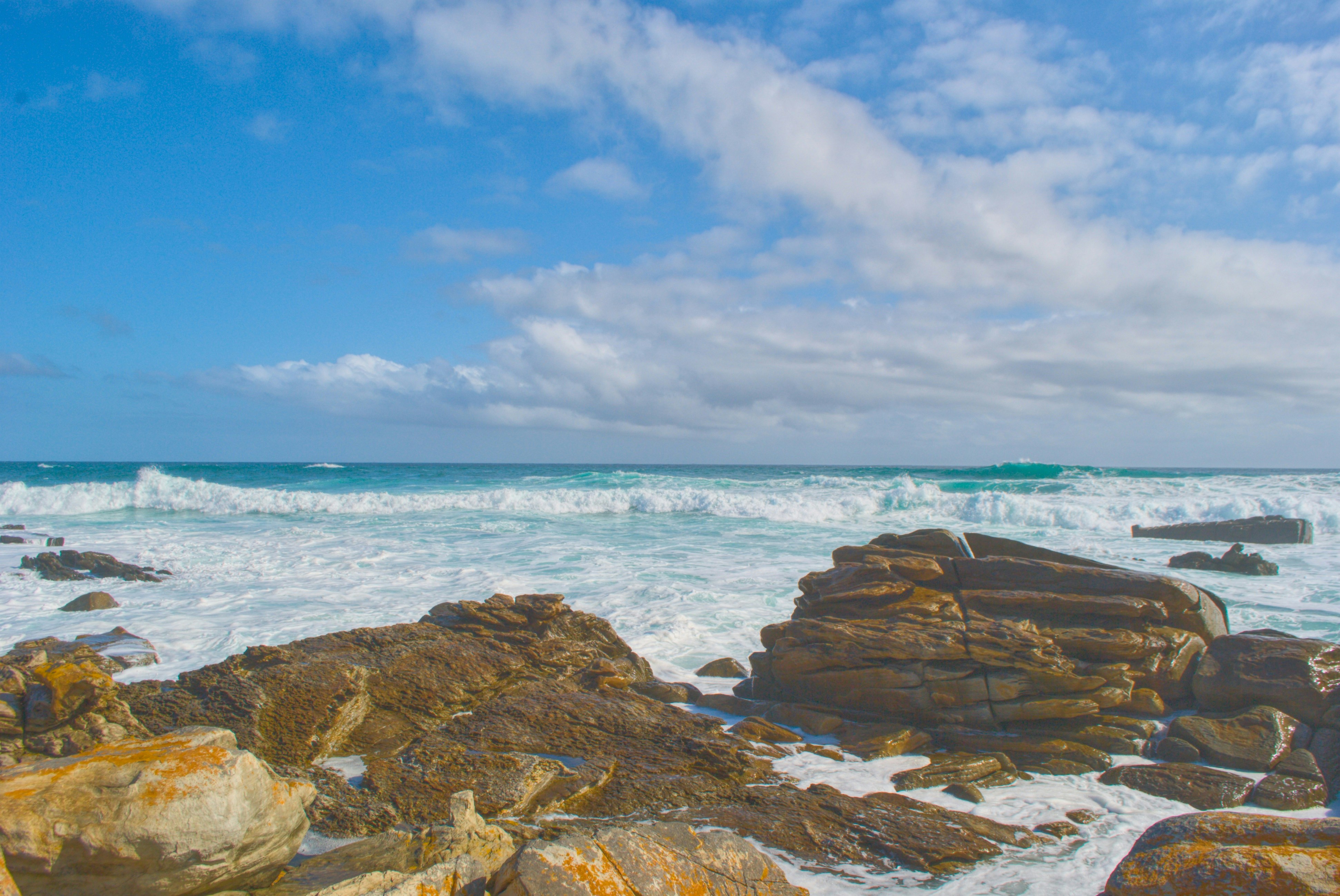 Ocean waves crash against jagged rocks under a blue sky with scattered clouds.