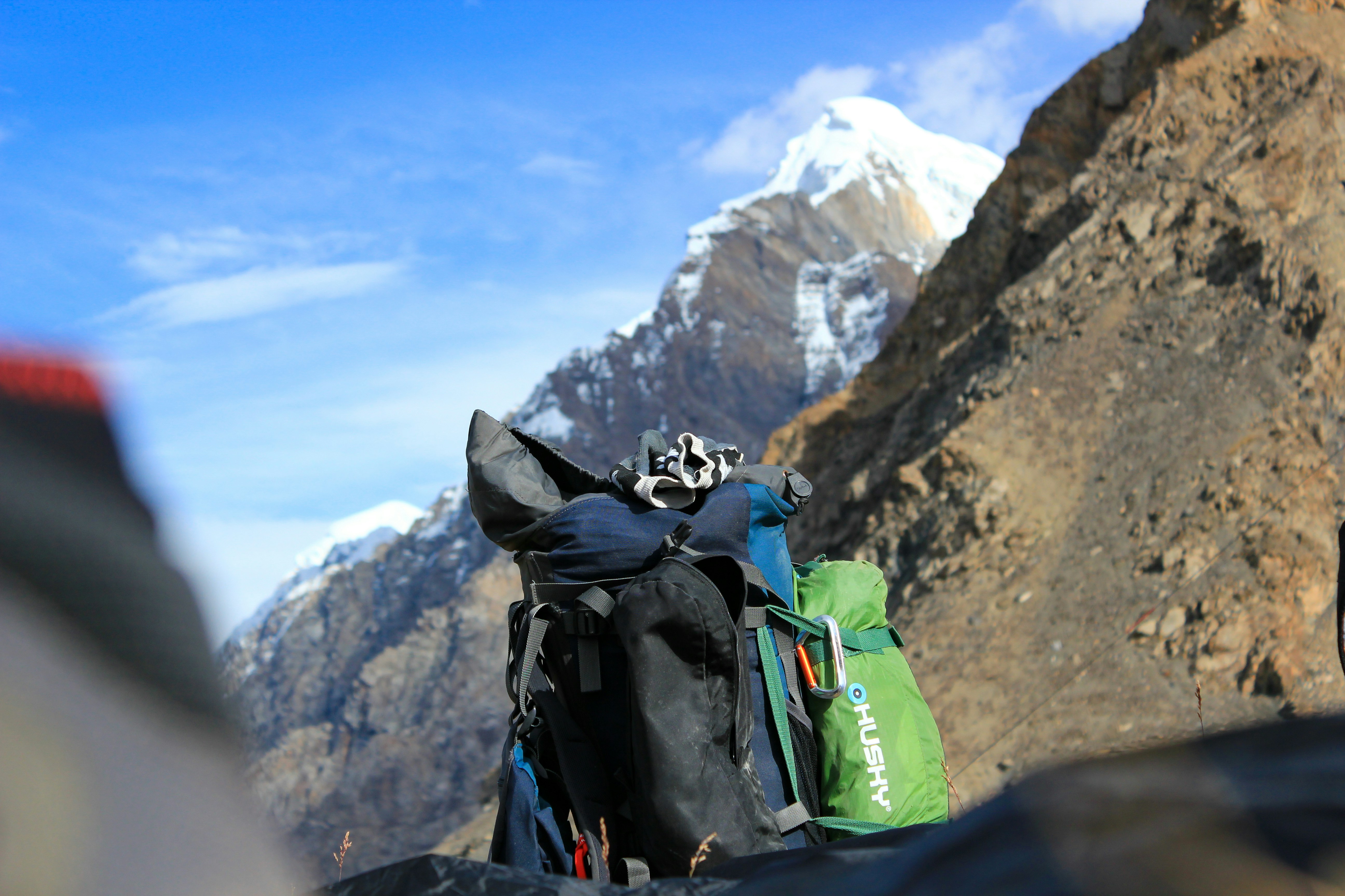 A well-worn backpack rests on rocky terrain, framed by towering snow-capped mountains under a clear blue sky.