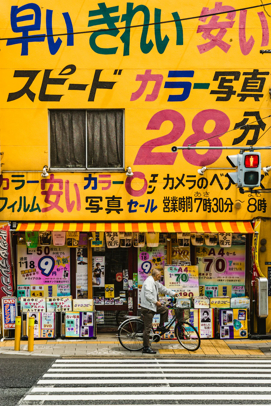 A vivid yellow Japanese storefront covered in large colorful signs and price boards, with a man on a bicycle passing in front. A crosswalk and traffic signal frame the busy street scene.