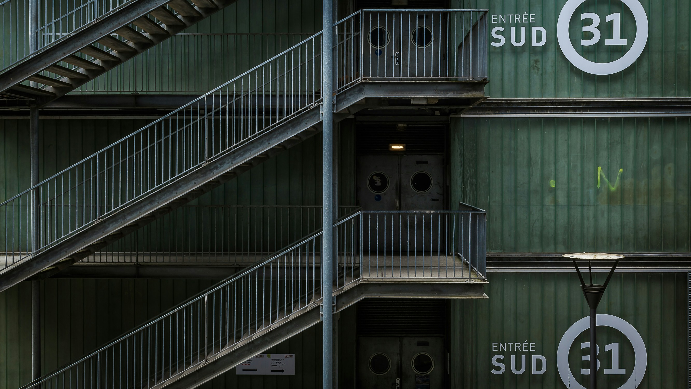 Metal staircases and green shipping container walls create a striking architectural composition, featuring bold signage. A street lamp adds a touch of urban ambiance.