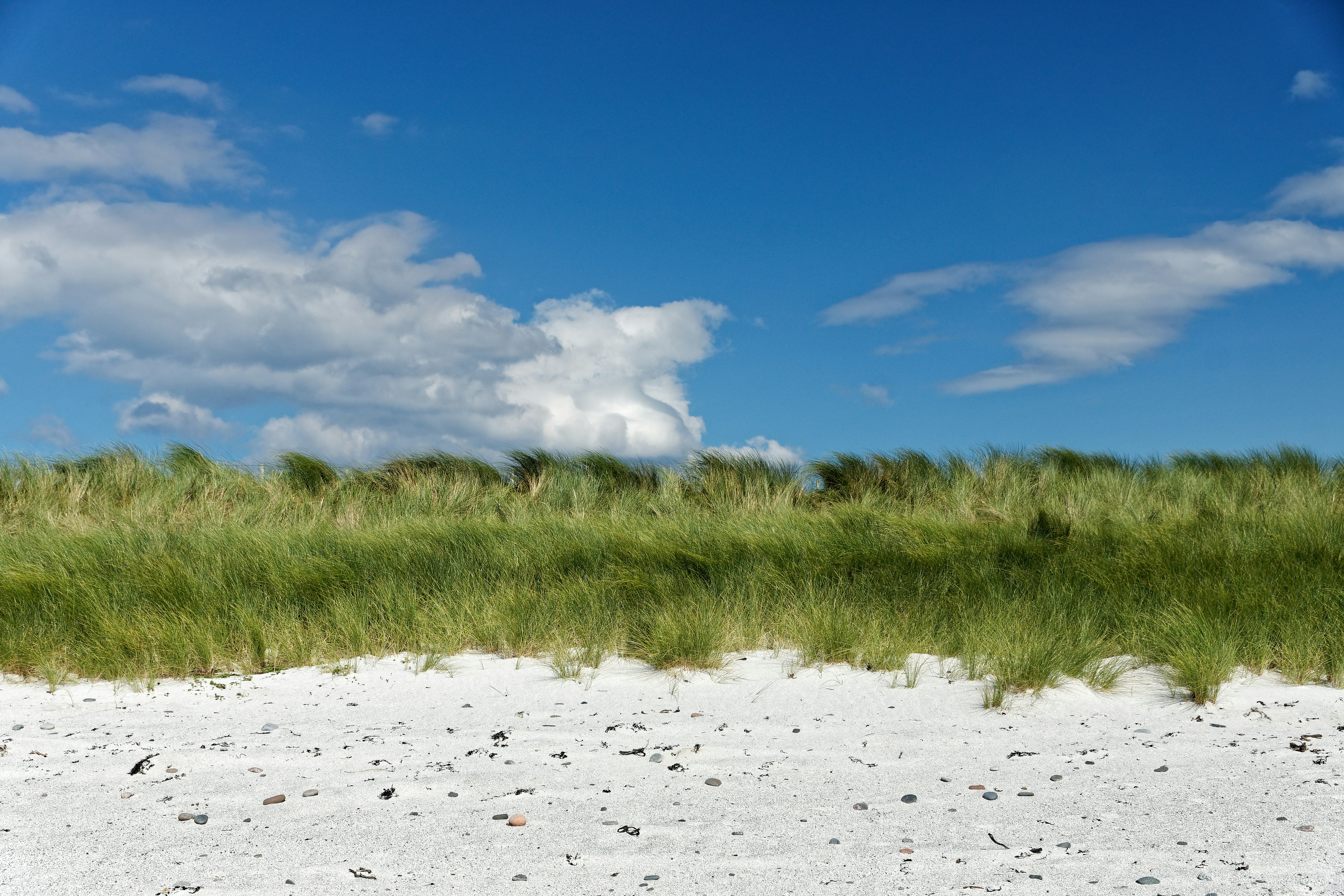 Sandy beach foreground with lush green grass and a vibrant blue sky filled with white clouds.
