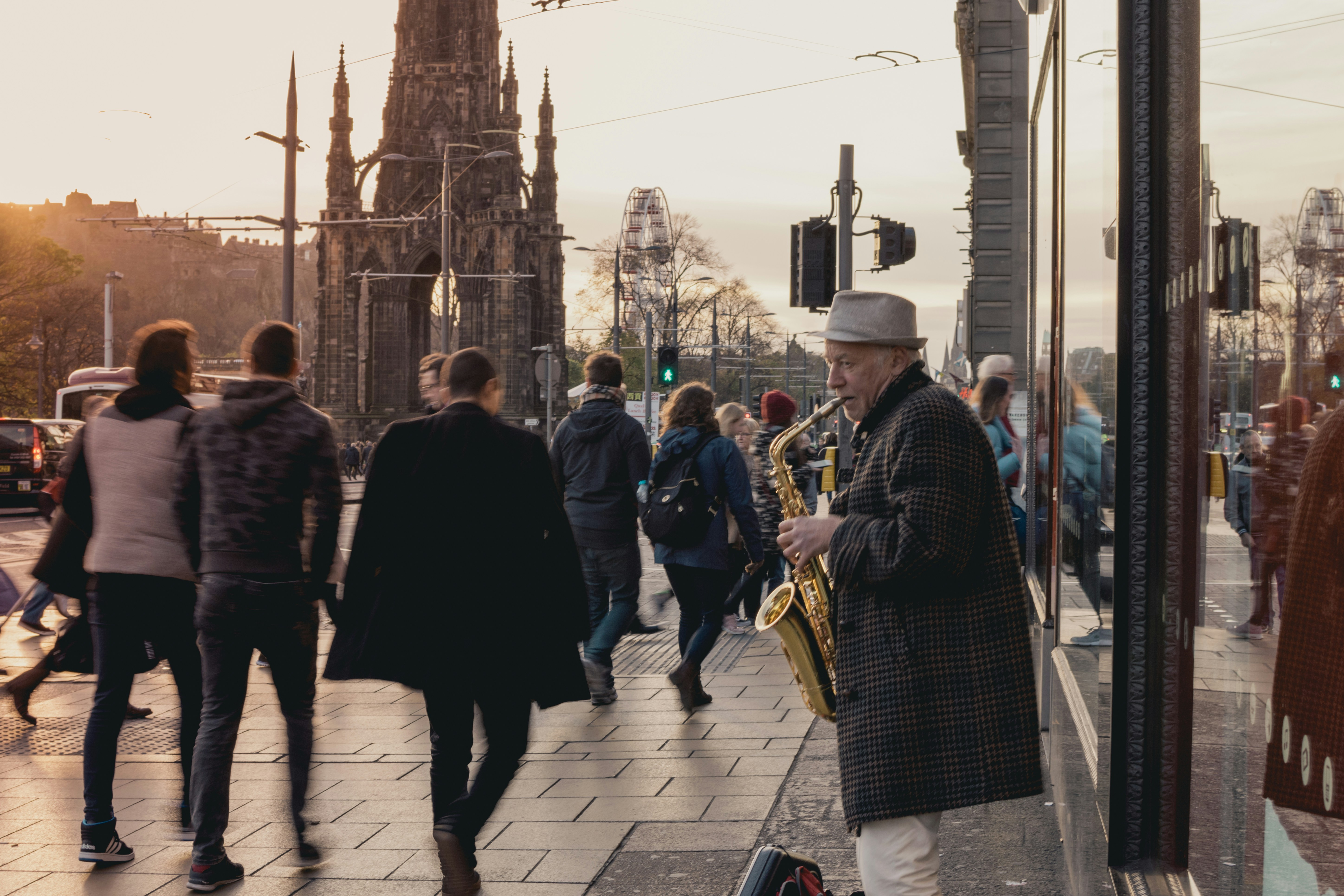 Street musician playing saxophone as pedestrians walk by during sunset.
