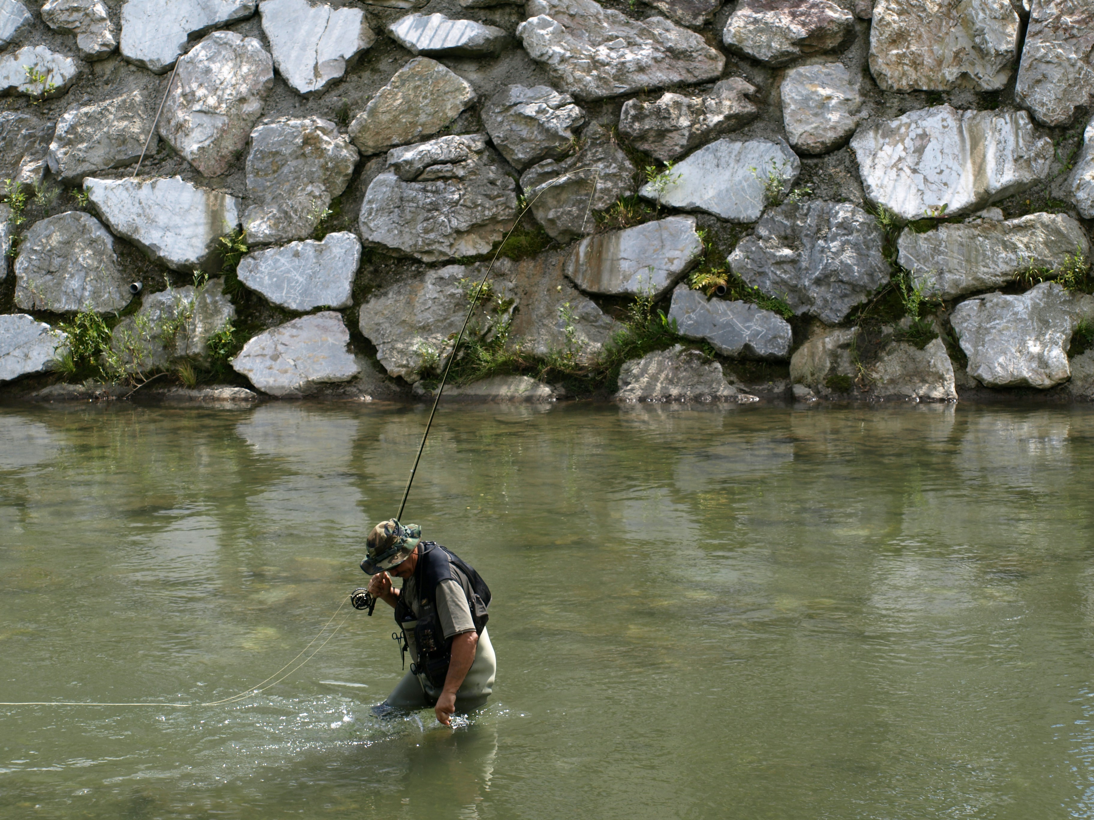 Person wading through a shallow, calm river against a backdrop of large stone wall.