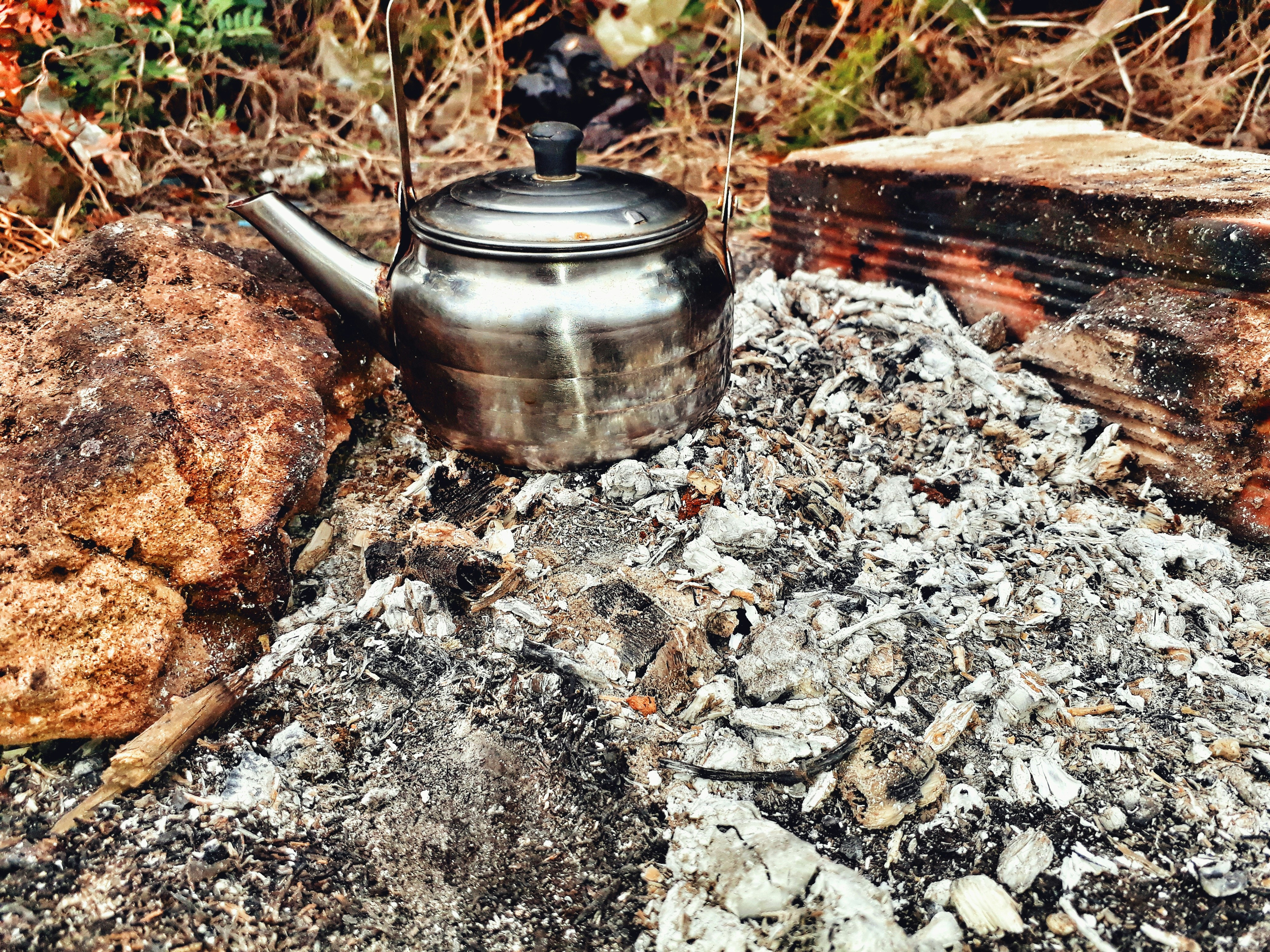 A metal teapot sits among embers and ashes, framed by rocks and a weathered log in an outdoor campfire scene.