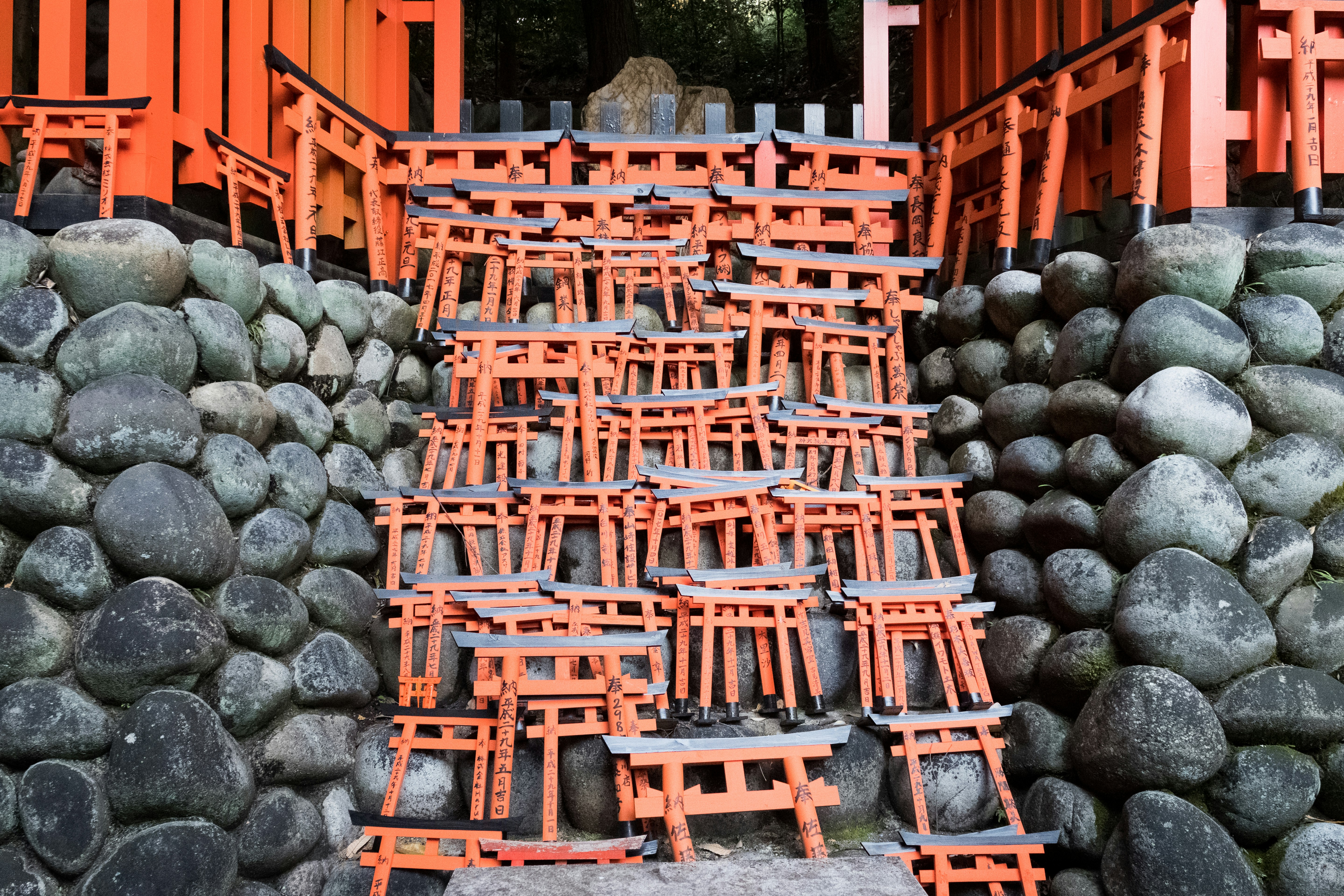 A cascade of miniature torii gates arranged on a stone staircase, surrounded by large rounded stones. The vibrant orange gates contrast with the natural stone texture.