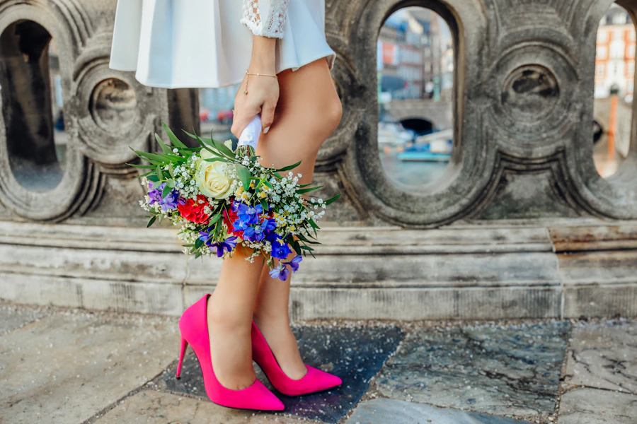 A woman in a white dress holds a colorful bouquet near her legs, wearing bright pink high heels beside an ornate stone balustrade.