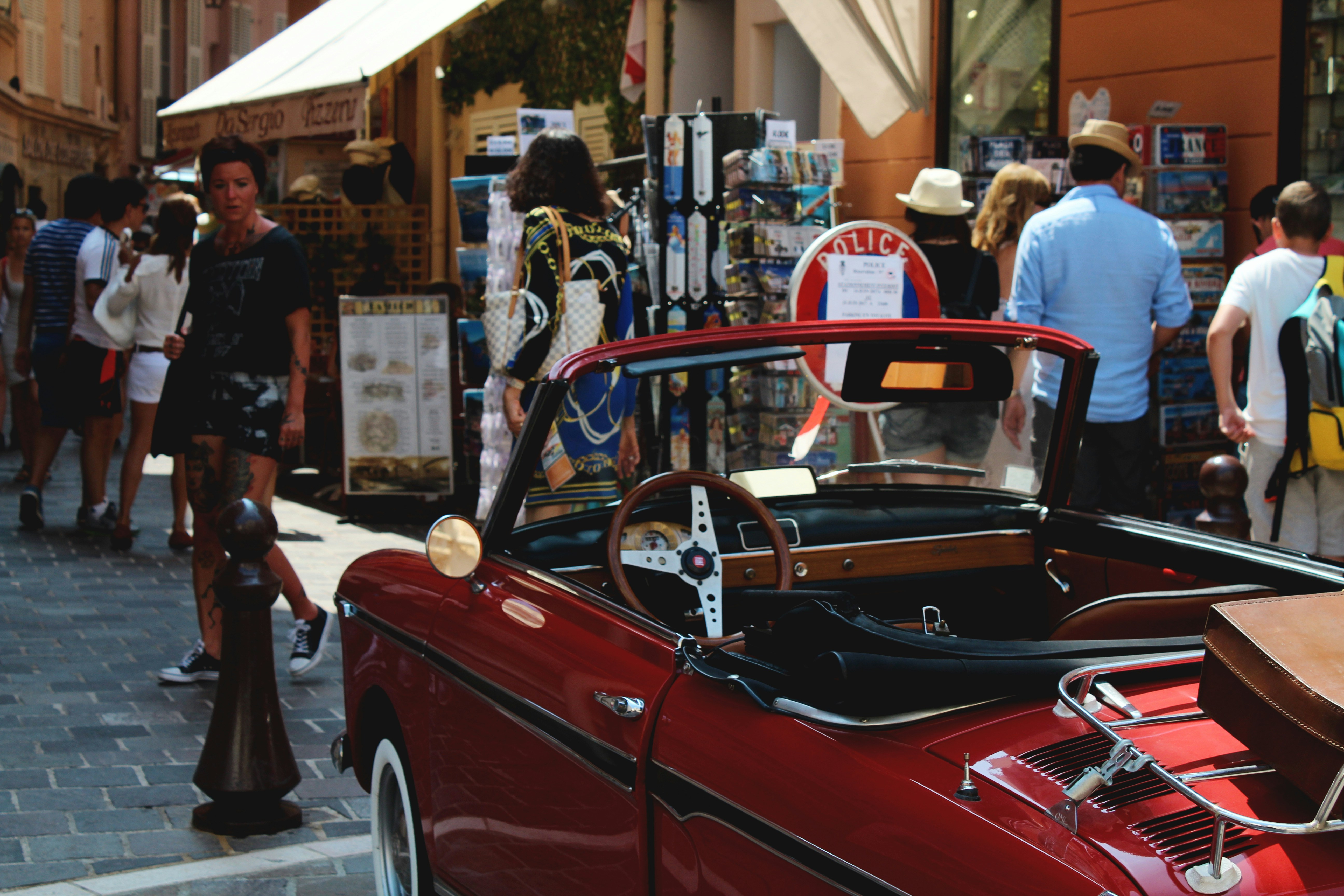 Red convertible parked near a bustling street market with people browsing shops.