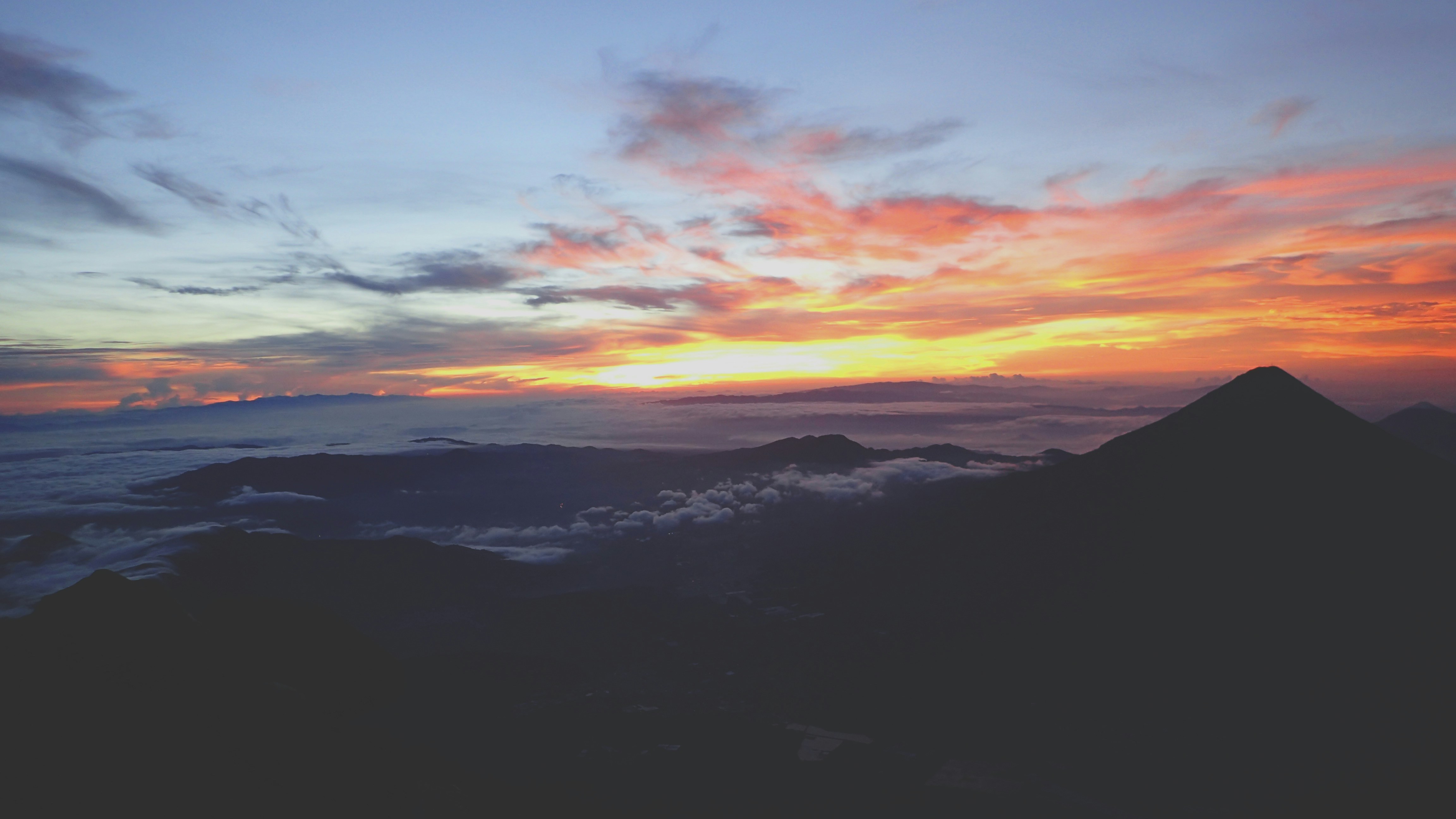 Silhouette of Volcan Fuego against a vibrant sunrise with clouds partially covering the city of Antigua.