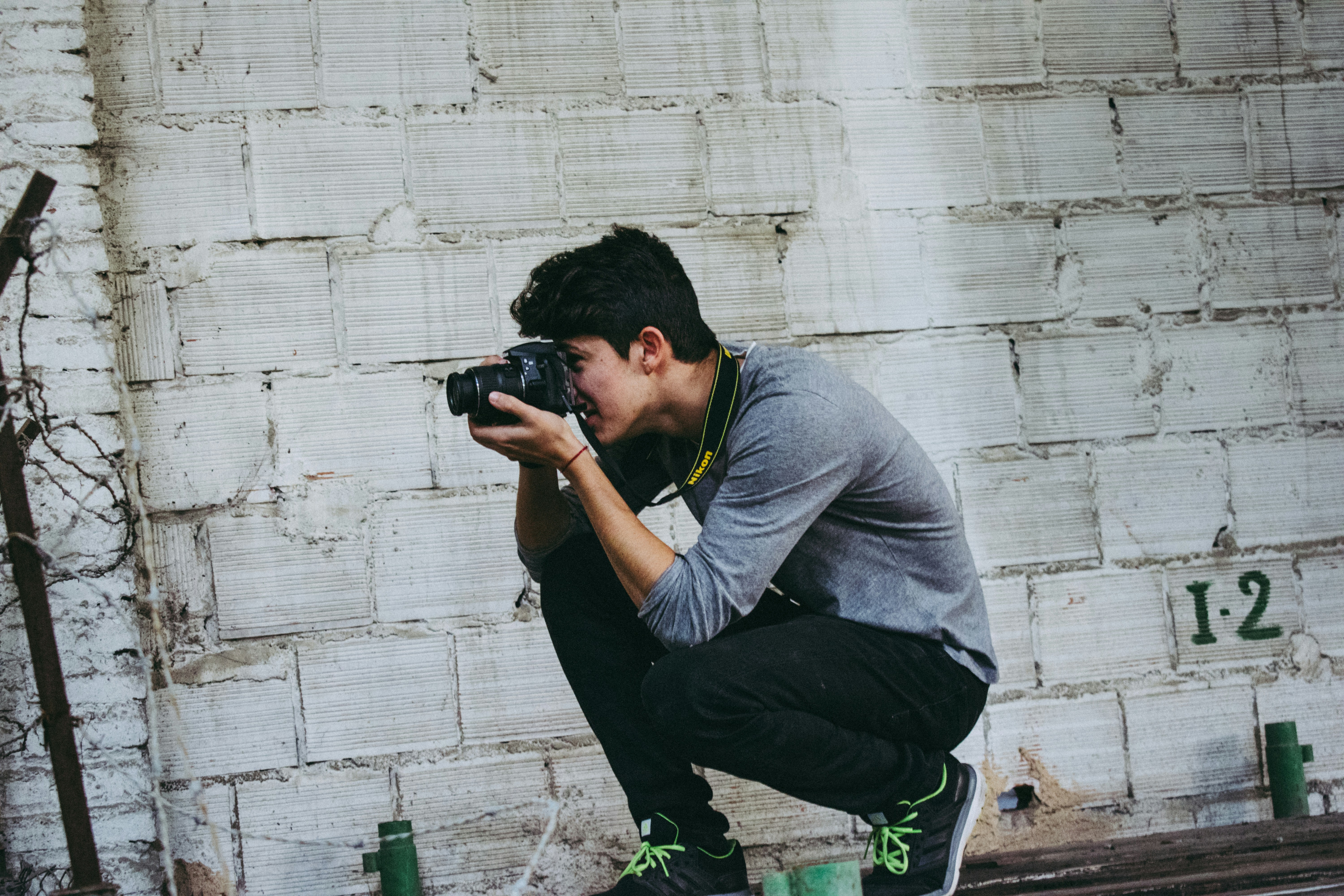 This candid shot features a young photographer crouched against a textured, industrial brick wall, absorbed in capturing the perfect frame with a Nikon camera. The scene's muted tones of gray and white contrast with the vivid green accents on the subject's shoes, creating a visually striking composition. The natural lighting casts soft shadows, adding depth and a contemplative mood to the image.