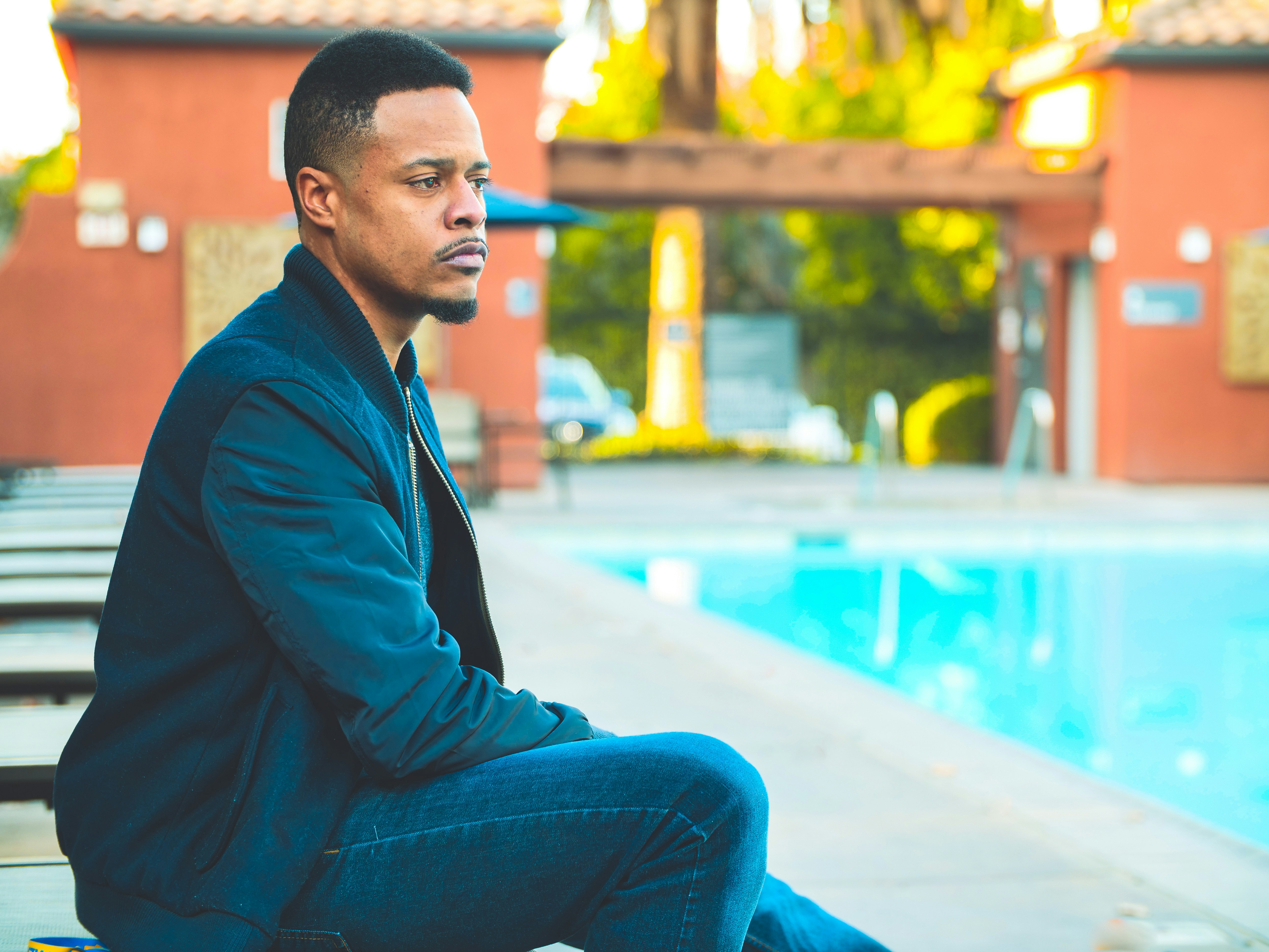 Man in dark-blue jacket seated near a tranquil pool with vibrant greenery and warm architecture in the background.