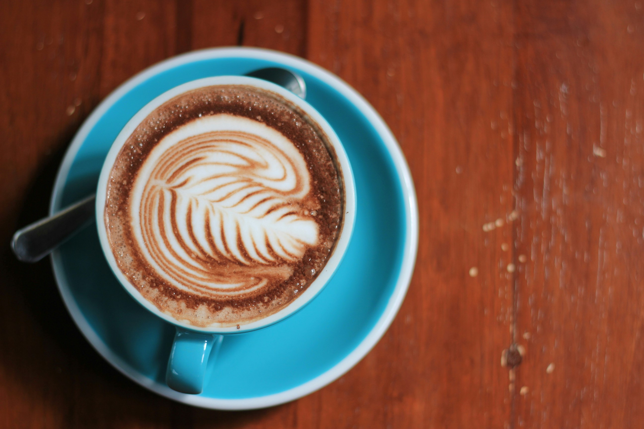 Artfully crafted cappuccino with a leaf design on frothy surface, set on a turquoise saucer against a rustic wooden table.