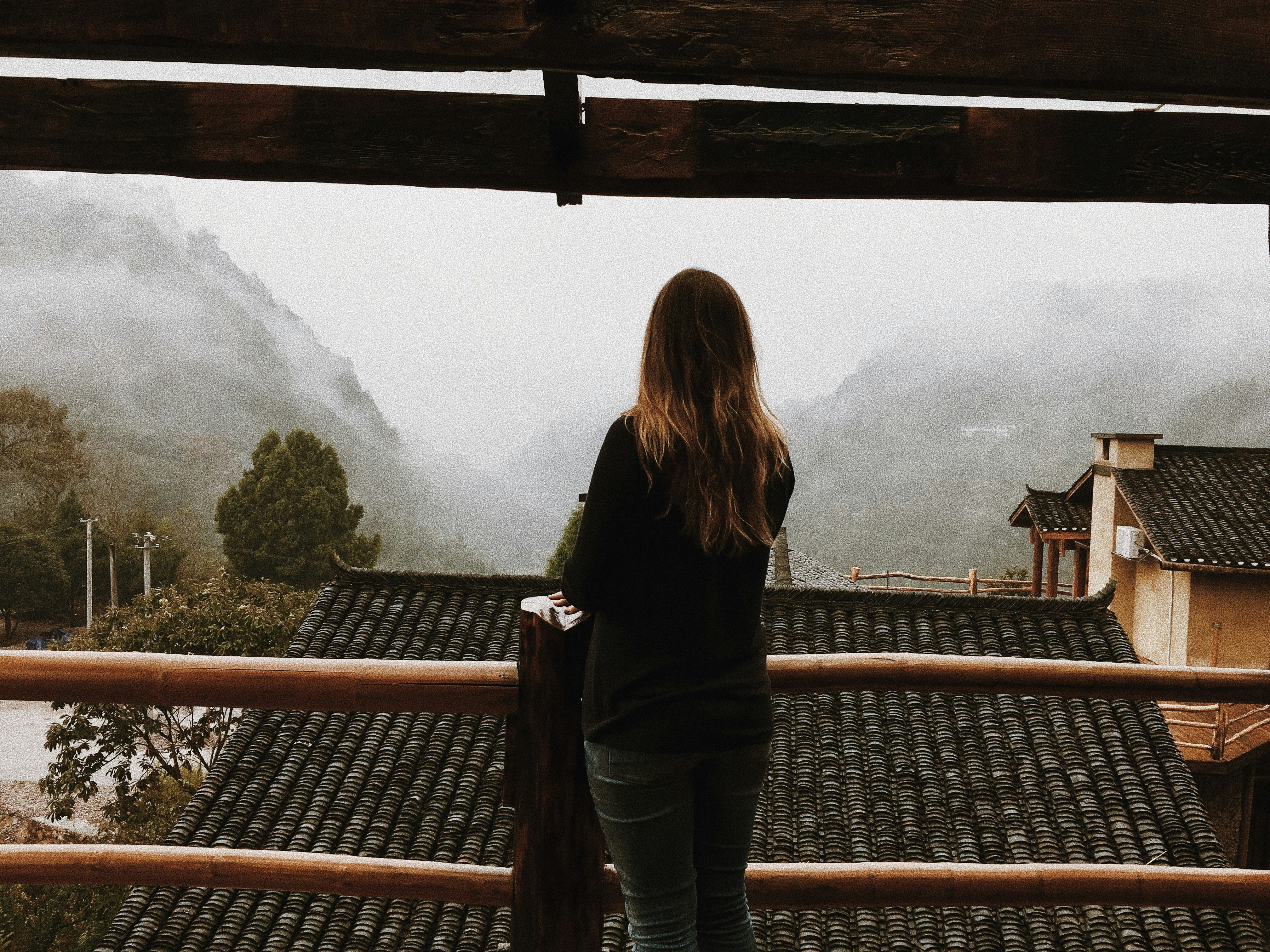 Person standing on a wooden balcony overlooking mist-covered mountains and traditional tiled rooftops.