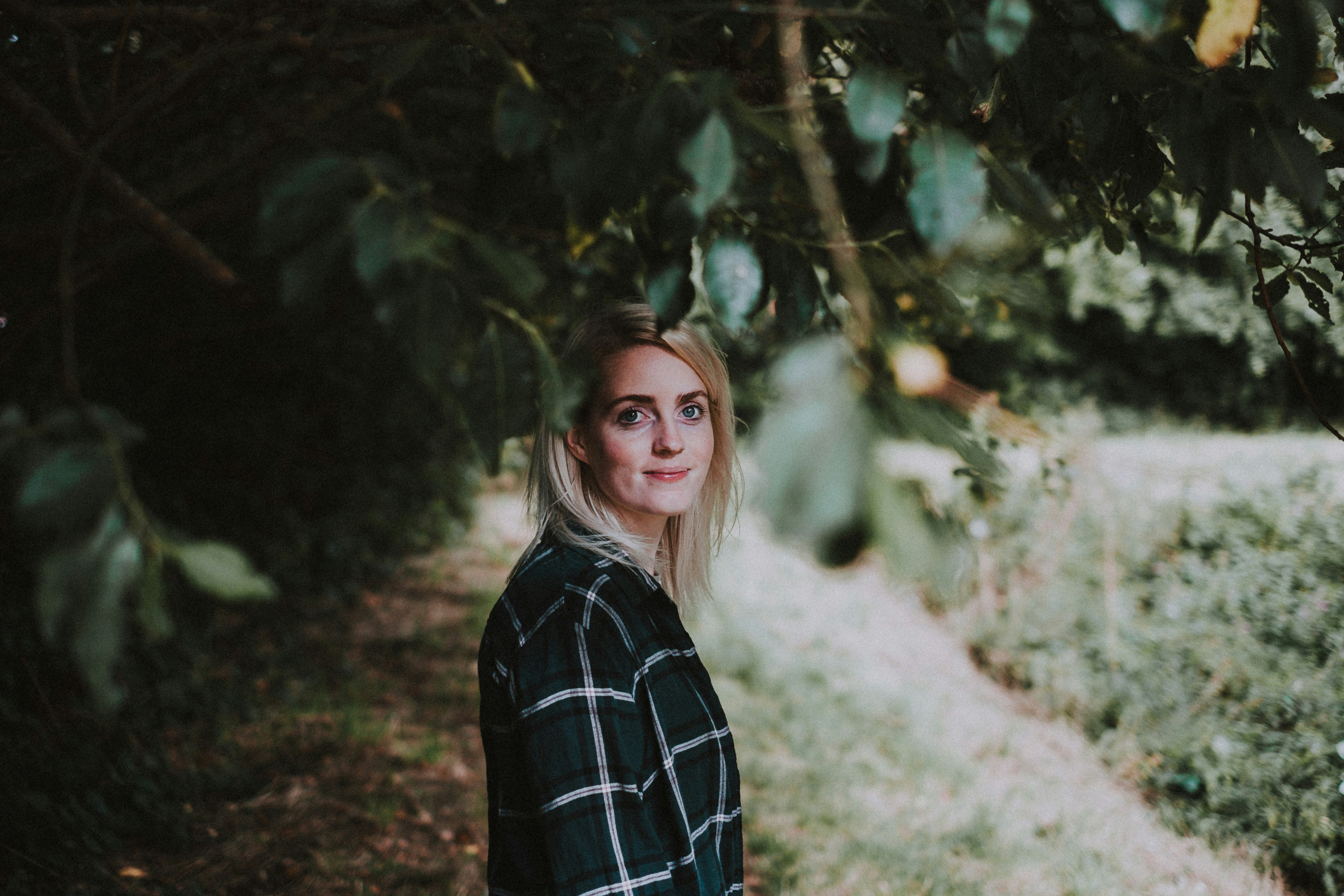 A woman stands amidst lush greenery, gazing thoughtfully while framed by overhanging leaves. The tranquil path behind her invites exploration.
