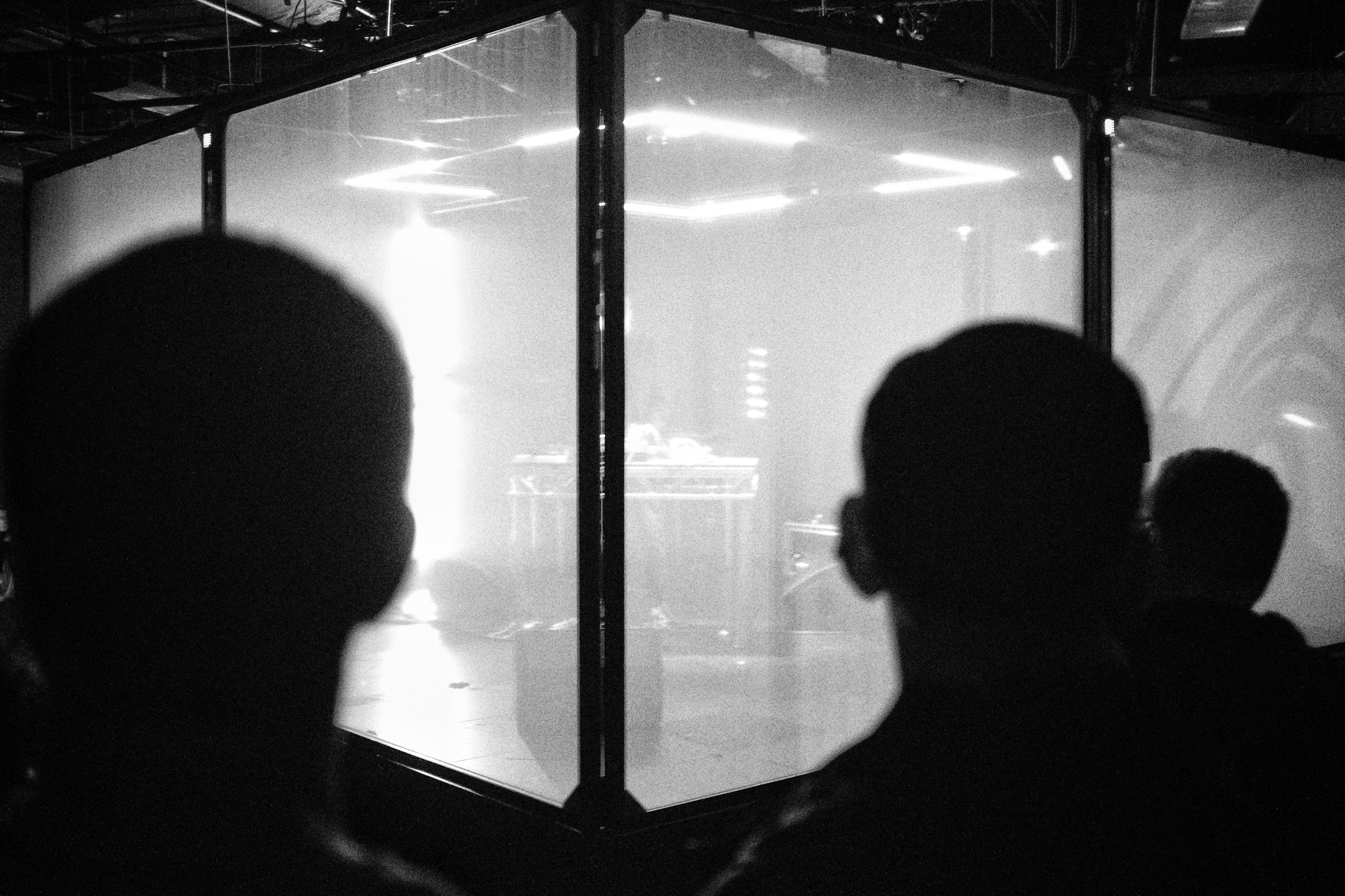 Silhouetted figures observe a glowing, misty glass enclosure in a dimly lit space.