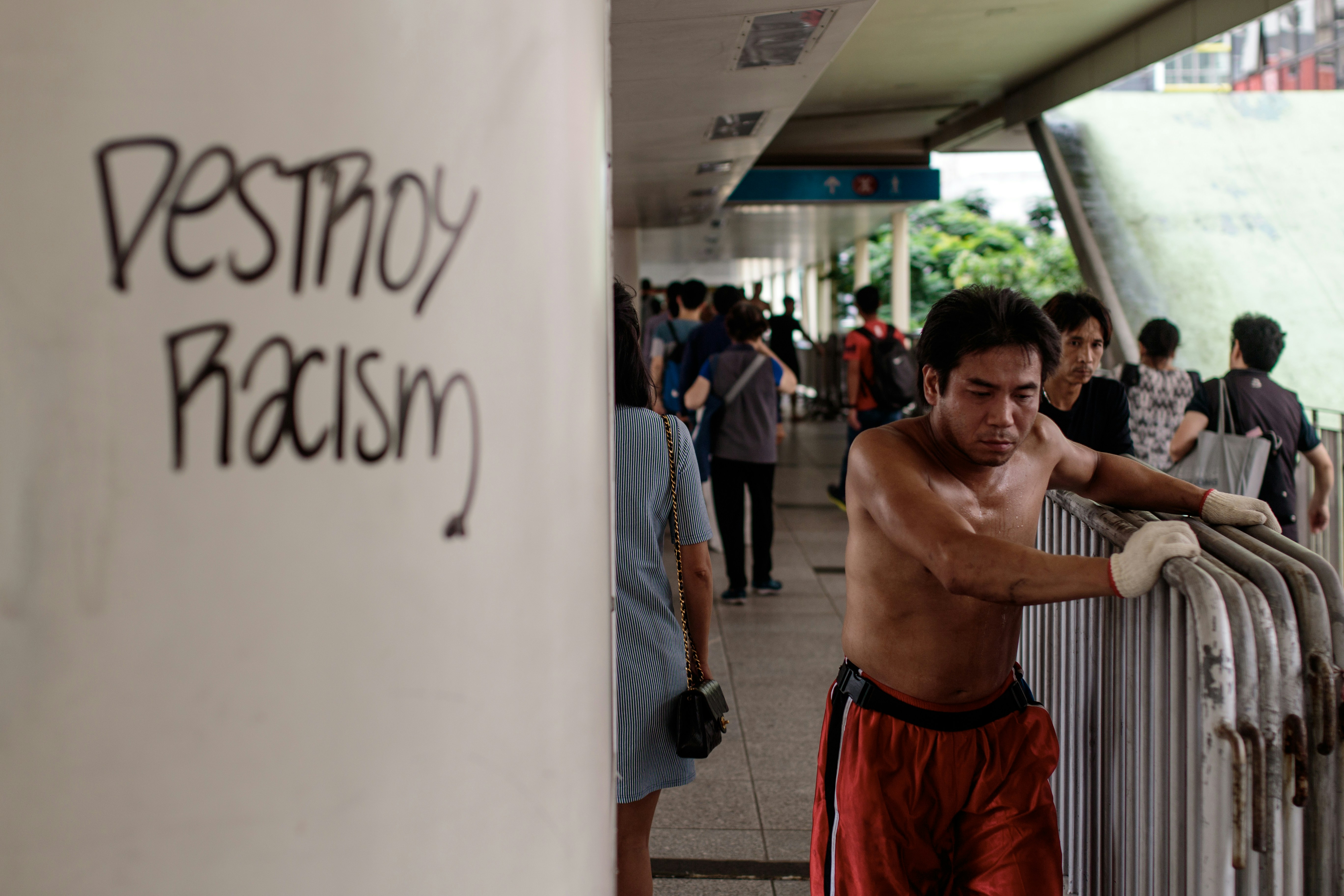 A shirtless man leans against a barrier, contemplating the message 'Destroy Racism' scrawled on the wall nearby. The scene captures a moment of reflection amidst a busy urban environment.