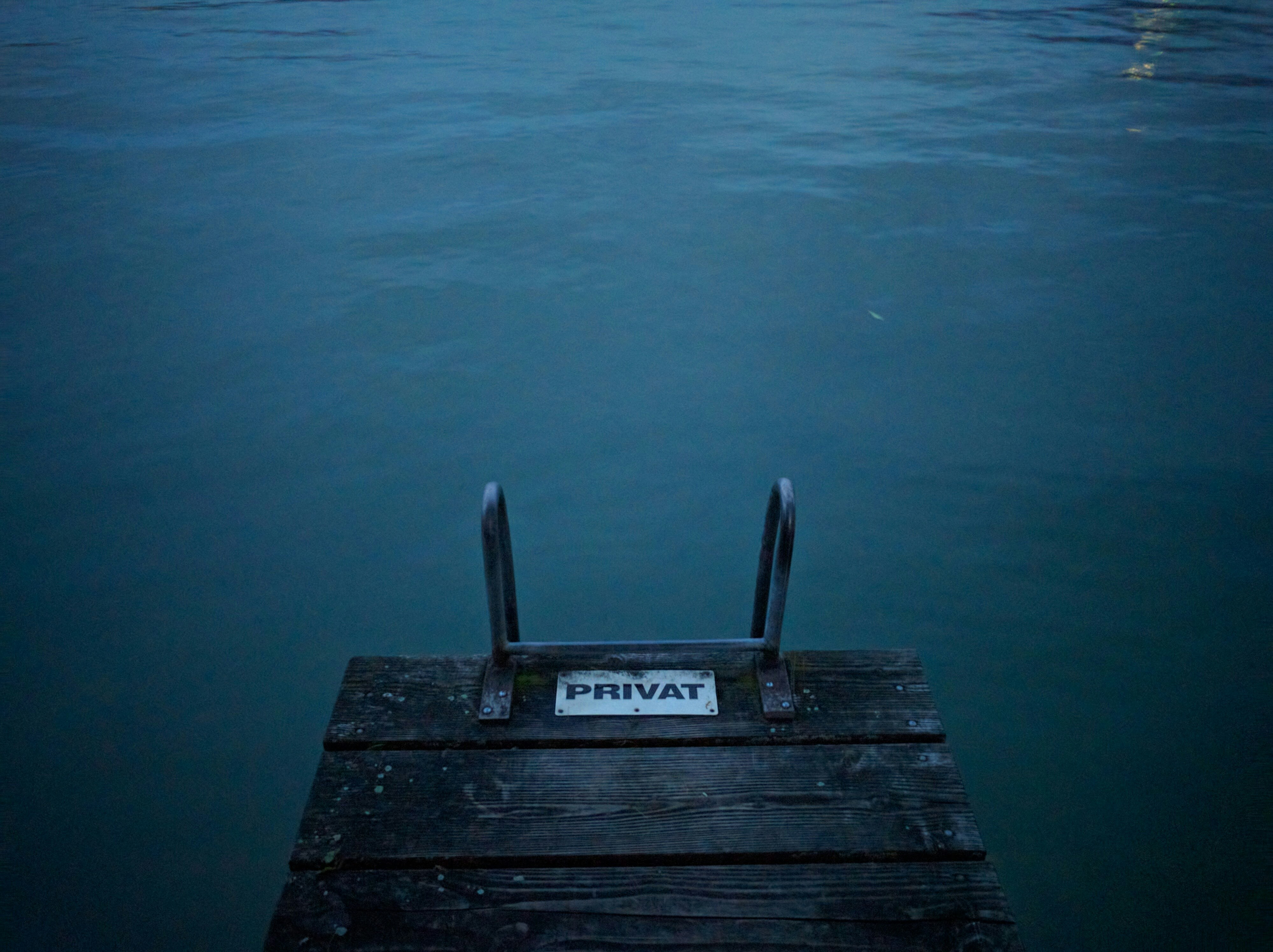 Photograph of a weathered wooden dock extending into calm blue water, with a small ladder and a 'PRIVAT' sign. The composition emphasizes solitude and moody blue tones.