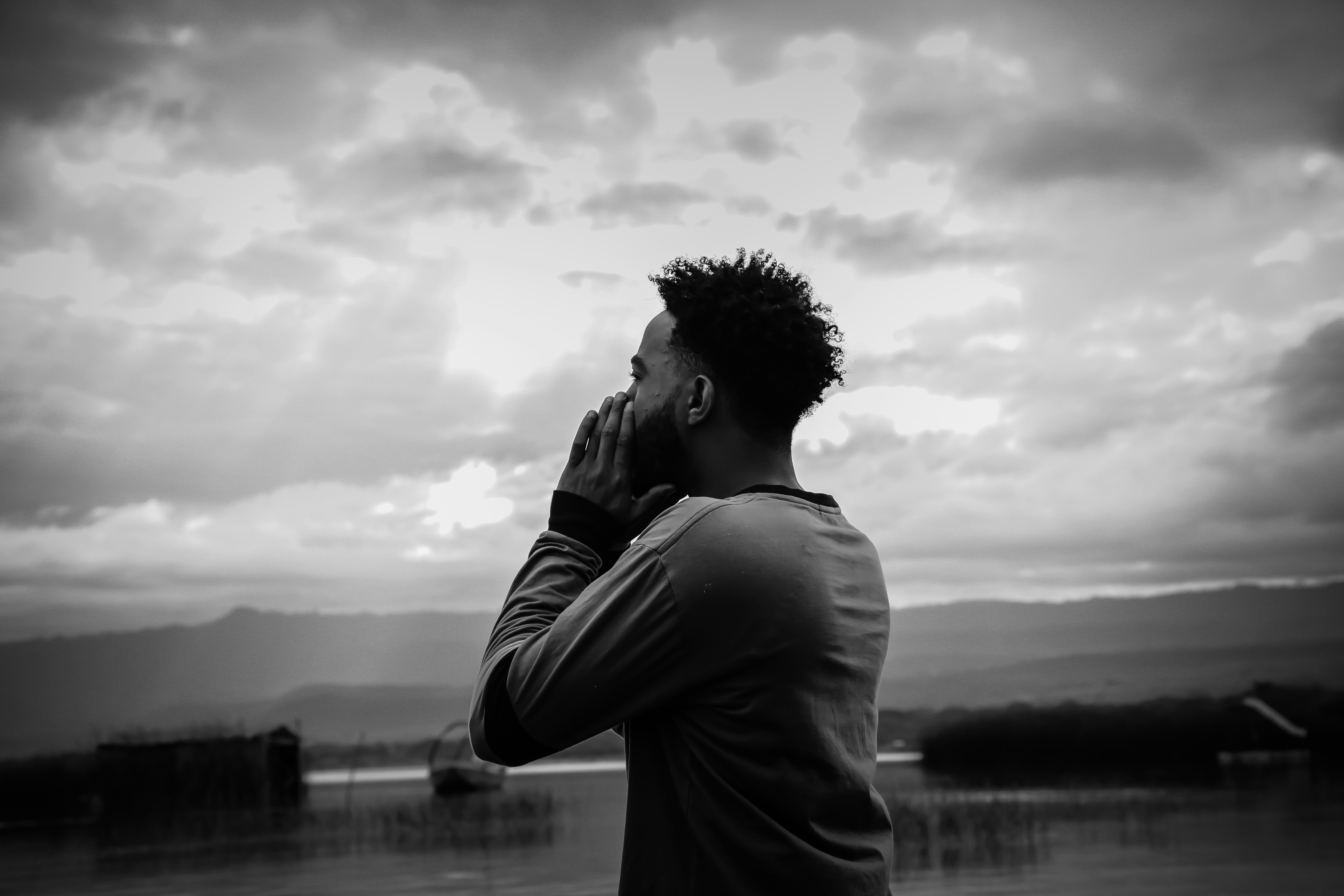 A person stands by a serene water body, calling out into the distance against a backdrop of cloudy skies. The monochrome tones enhance the emotional depth of the scene.