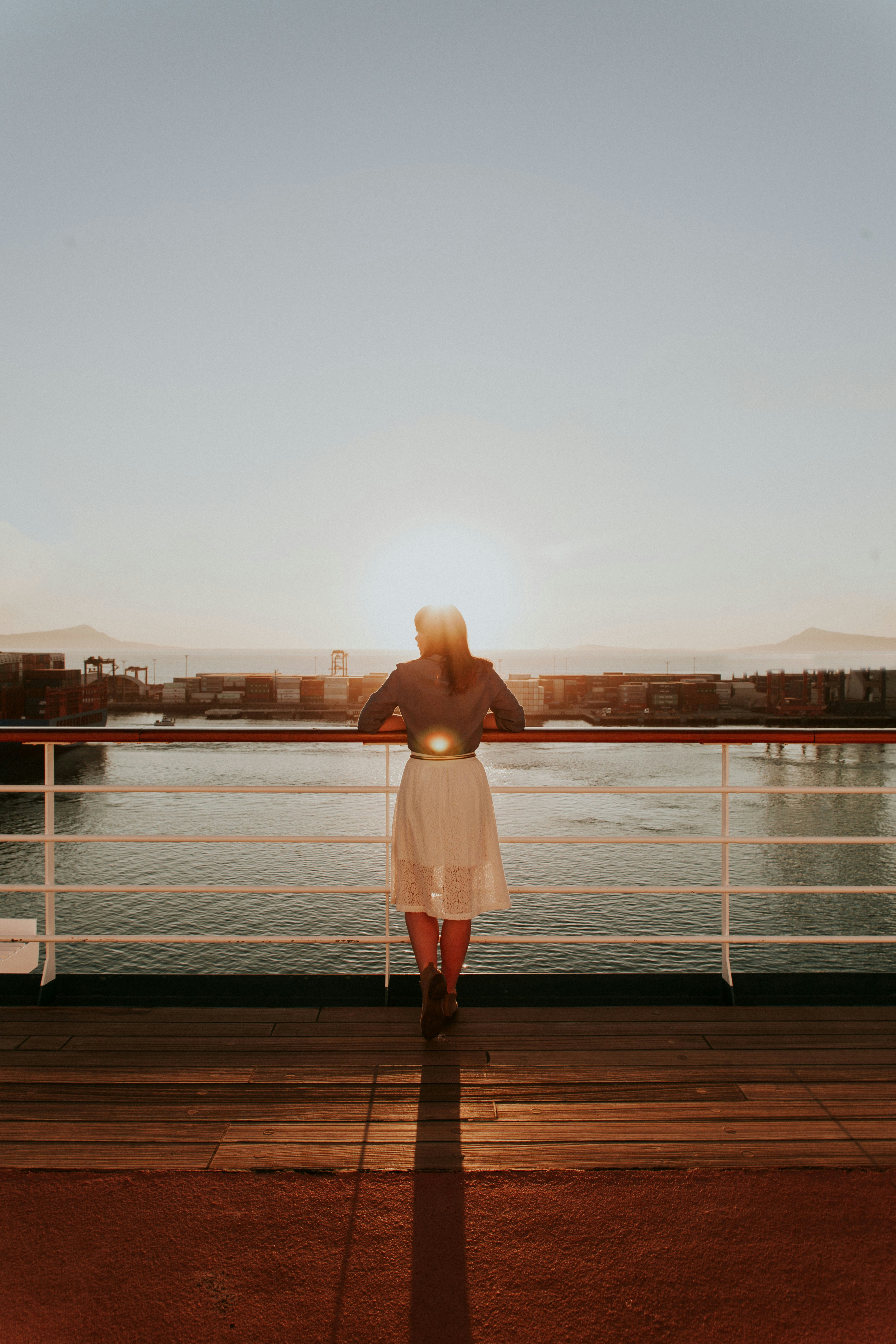 Woman in a white skirt gazing over a ship's railing at a sunlit harbor.