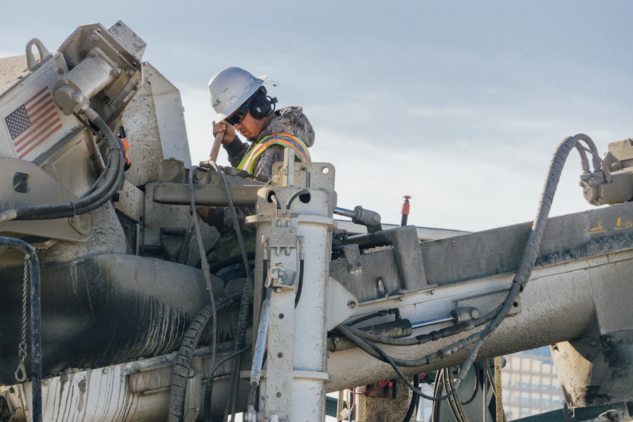 Construction worker in a white hard hat inspects exposed piping and metal framework atop heavy industrial machinery. The shot highlights field maintenance in a high-workload industrial environment.