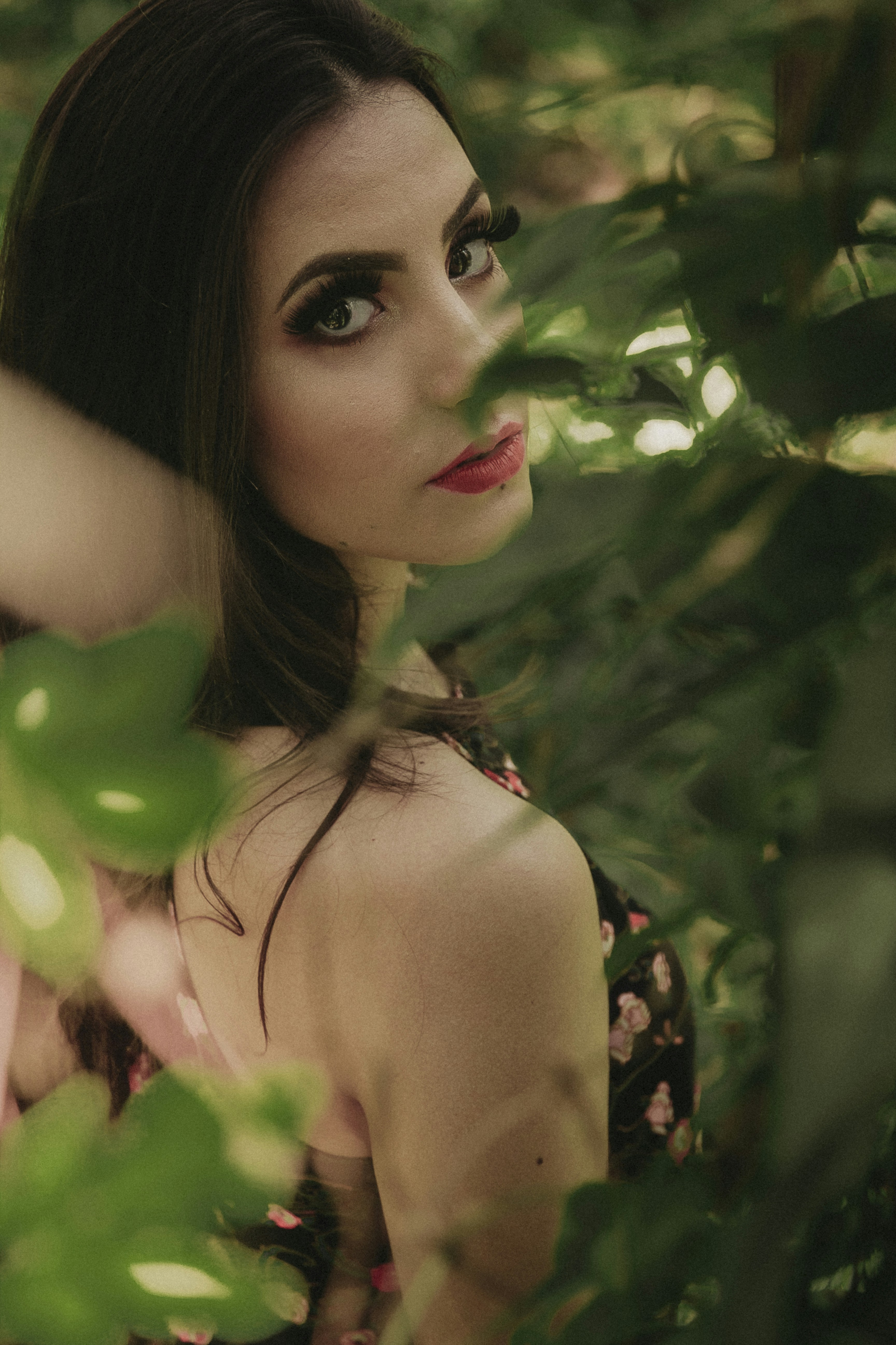 A woman gazes thoughtfully through lush foliage, her striking features framed by vibrant green leaves. The interplay of light and shadow enhances the intimate atmosphere.