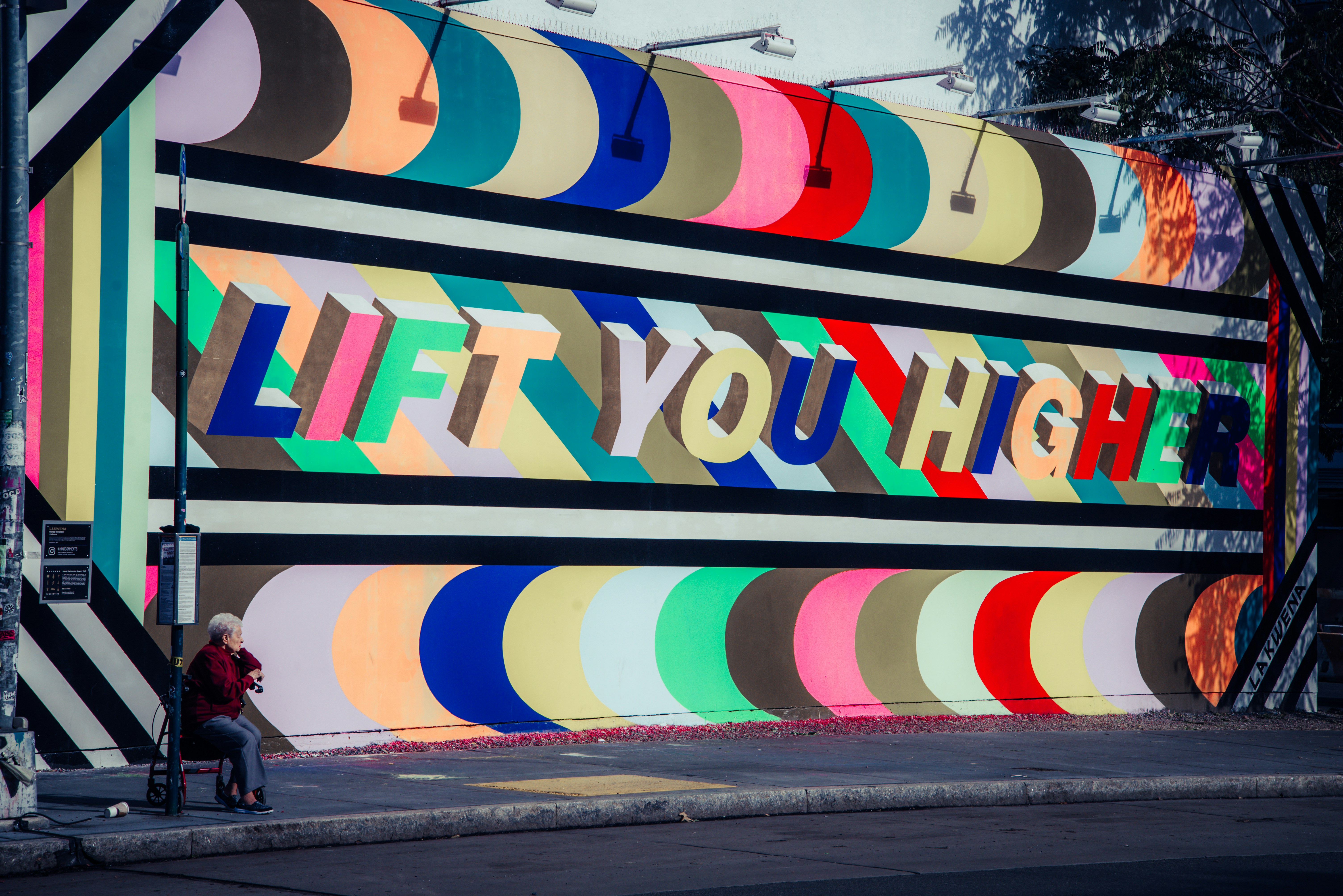 Person seated in front of a colorful mural with the words 'Lift You Higher.'.