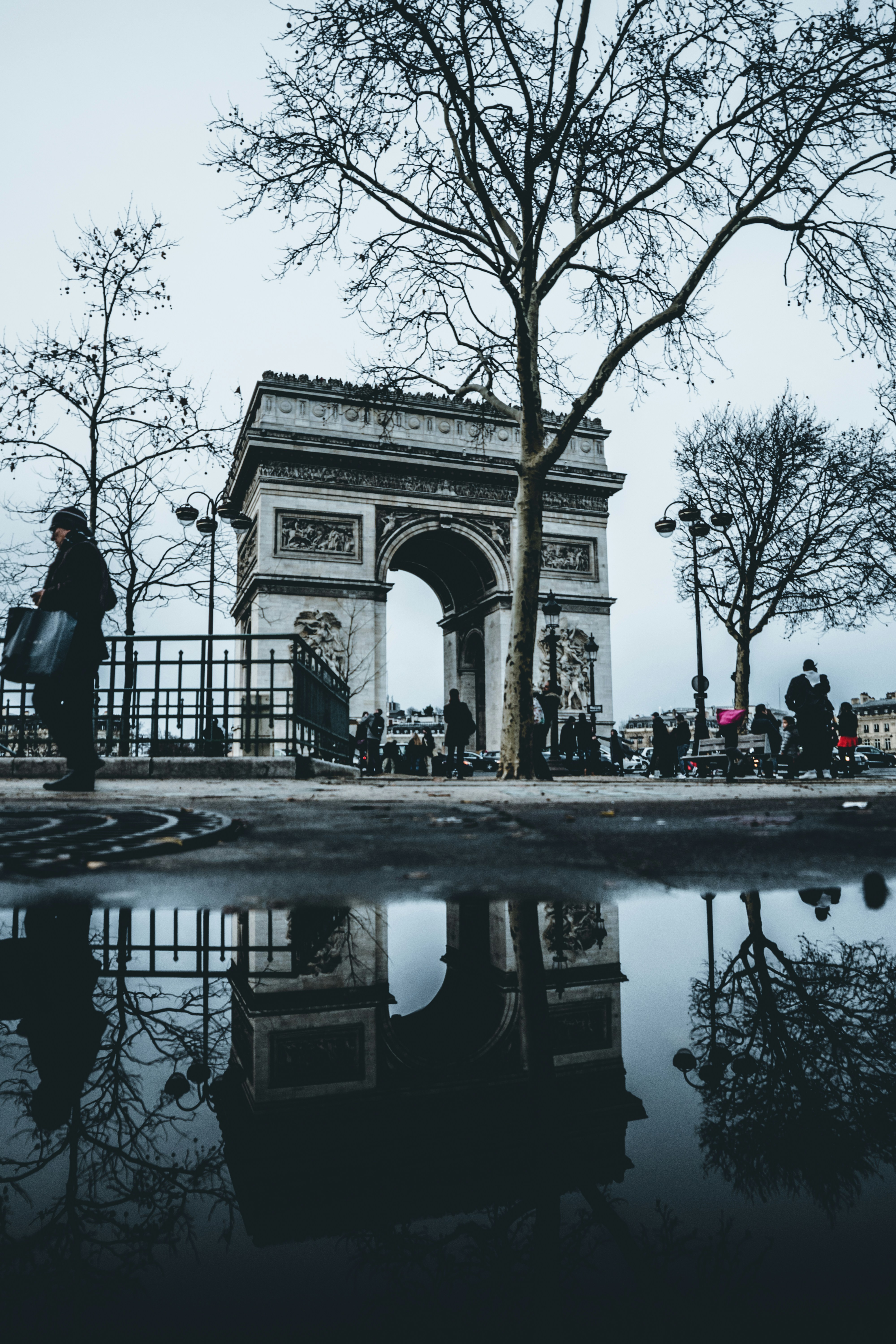 The Arc de Triomphe stands majestically as its reflection shimmers in a puddle, surrounded by bare trees and bustling pedestrians.