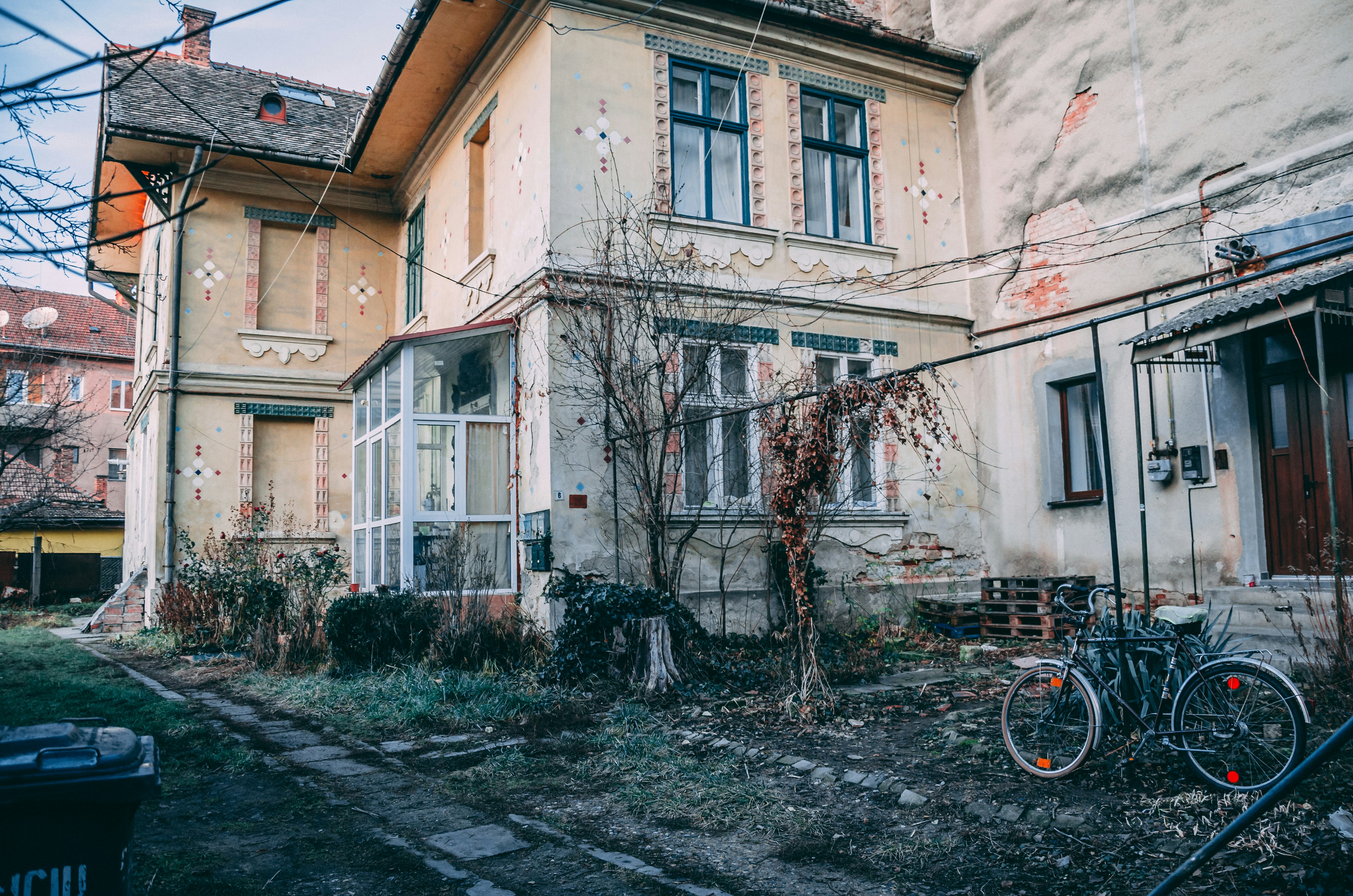 A weathered building with peeling paint and overgrown vegetation, featuring a glass-enclosed porch and a vintage bicycle parked nearby.
