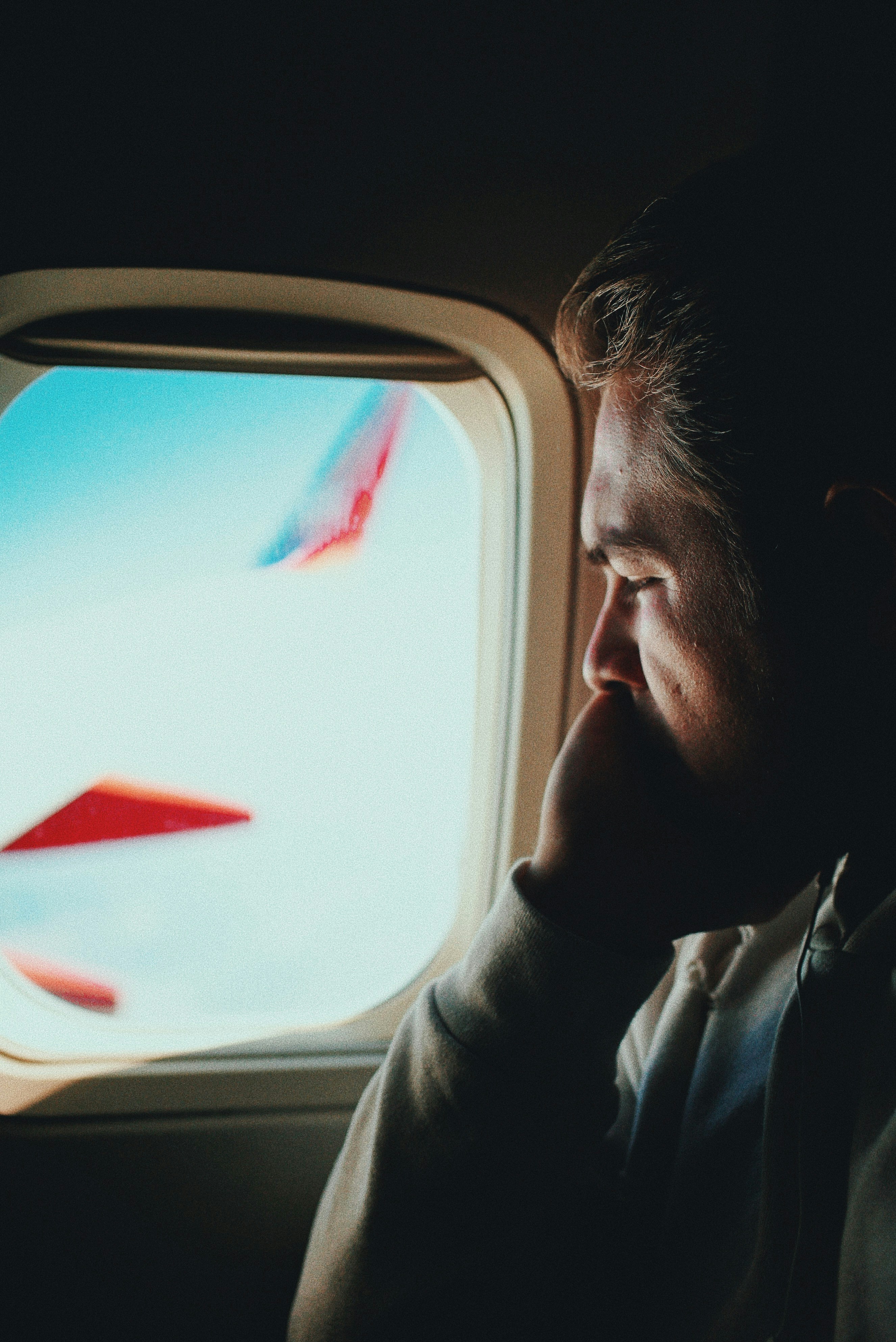 A contemplative traveler gazes out of an airplane window, with the wing and bright sky in the background. The scene captures a moment of introspection during flight.