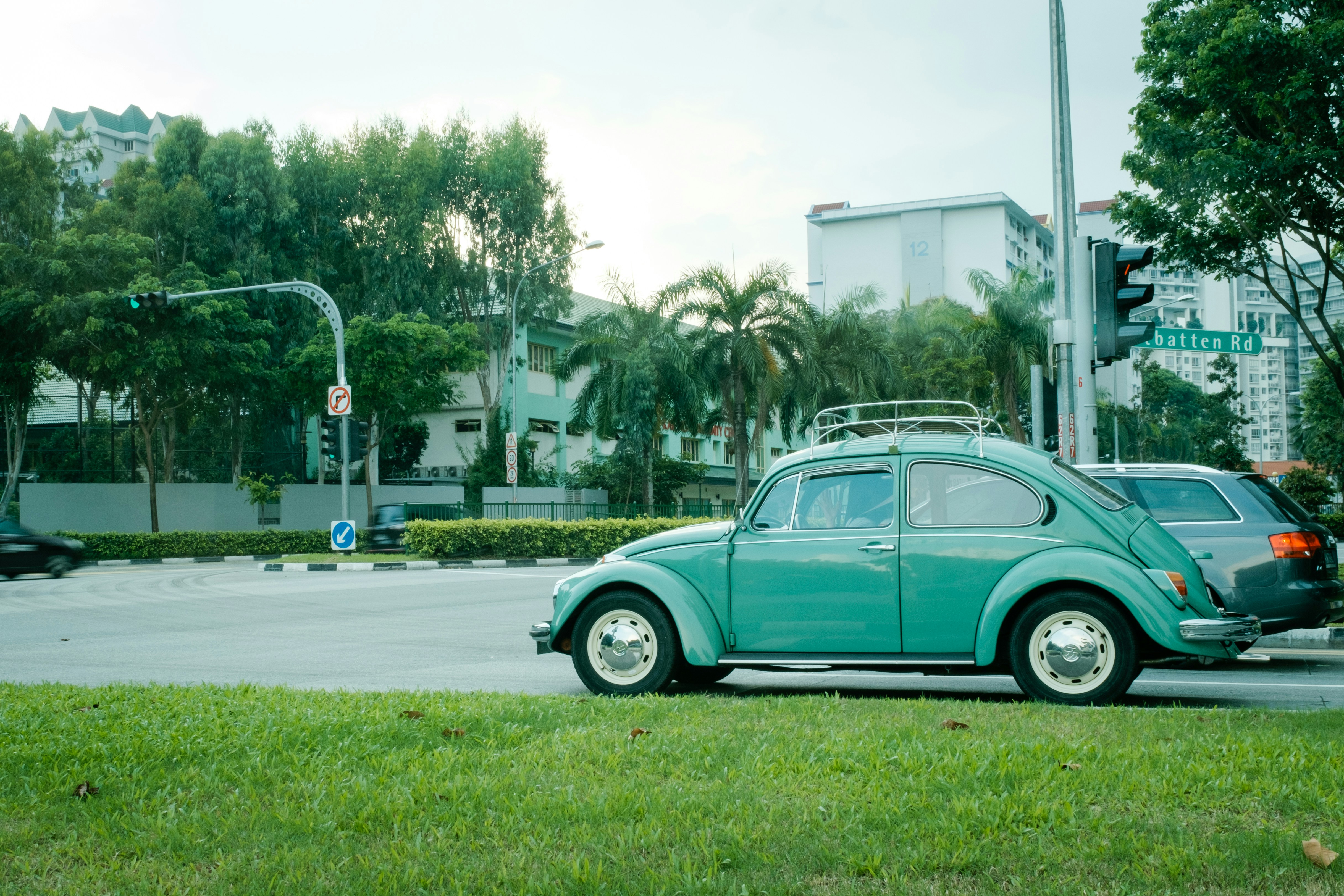 A vintage green Volkswagen Beetle parked beside a busy intersection, surrounded by lush greenery and modern buildings.