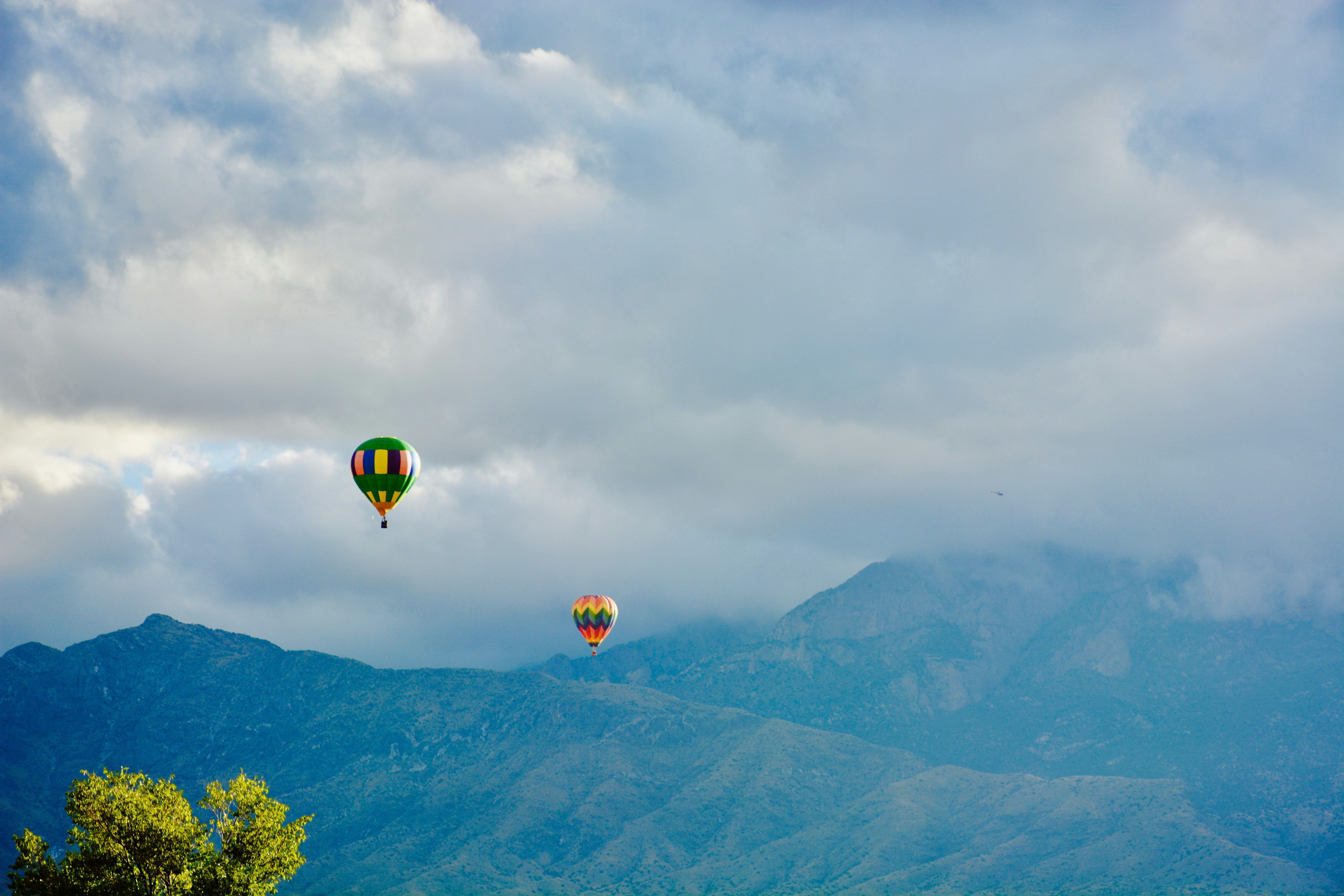 Two colorful hot air balloons drift above a mountain range under a cloudy sky.