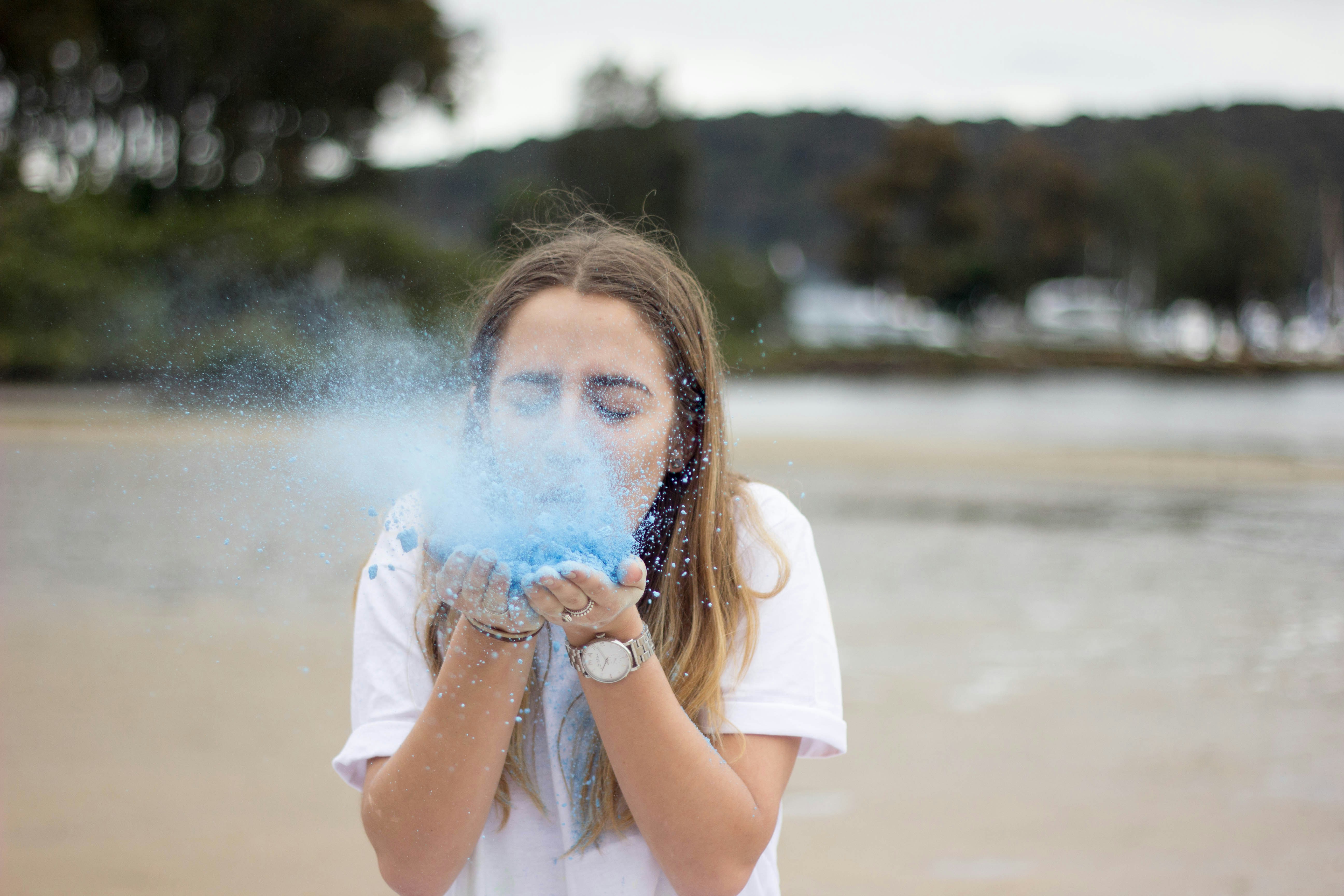 Young woman playfully blowing blue powder into the air on a sandy beach, with a blurred natural background. The scene captures a moment of joy and creativity.