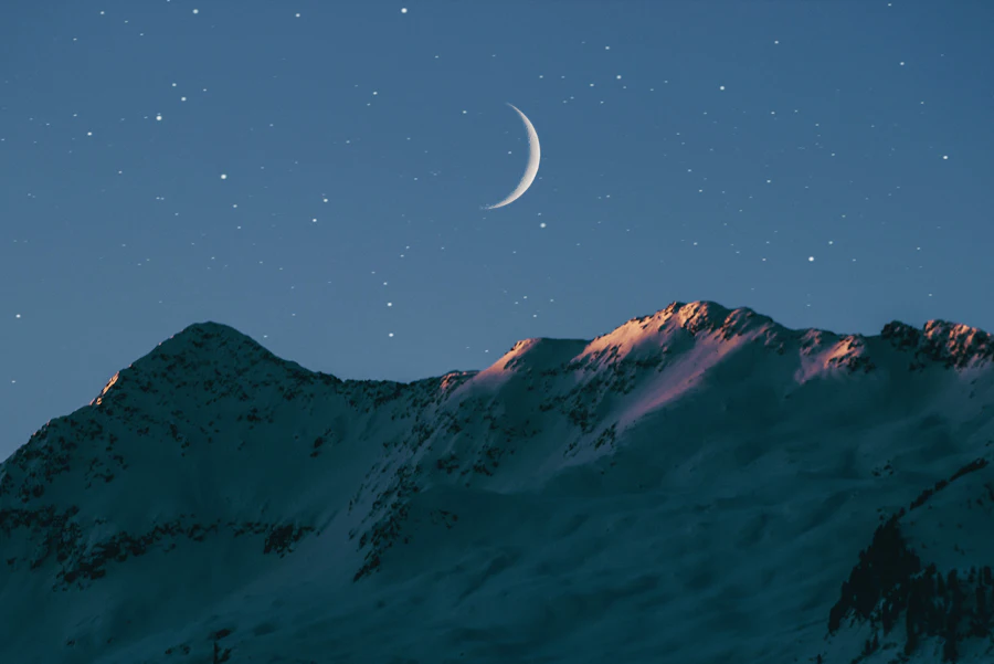 Thin crescent moon hangs above a range of snow-covered mountains under a deep blue night sky. Pink light catches the ridge line while scattered stars dot the background.