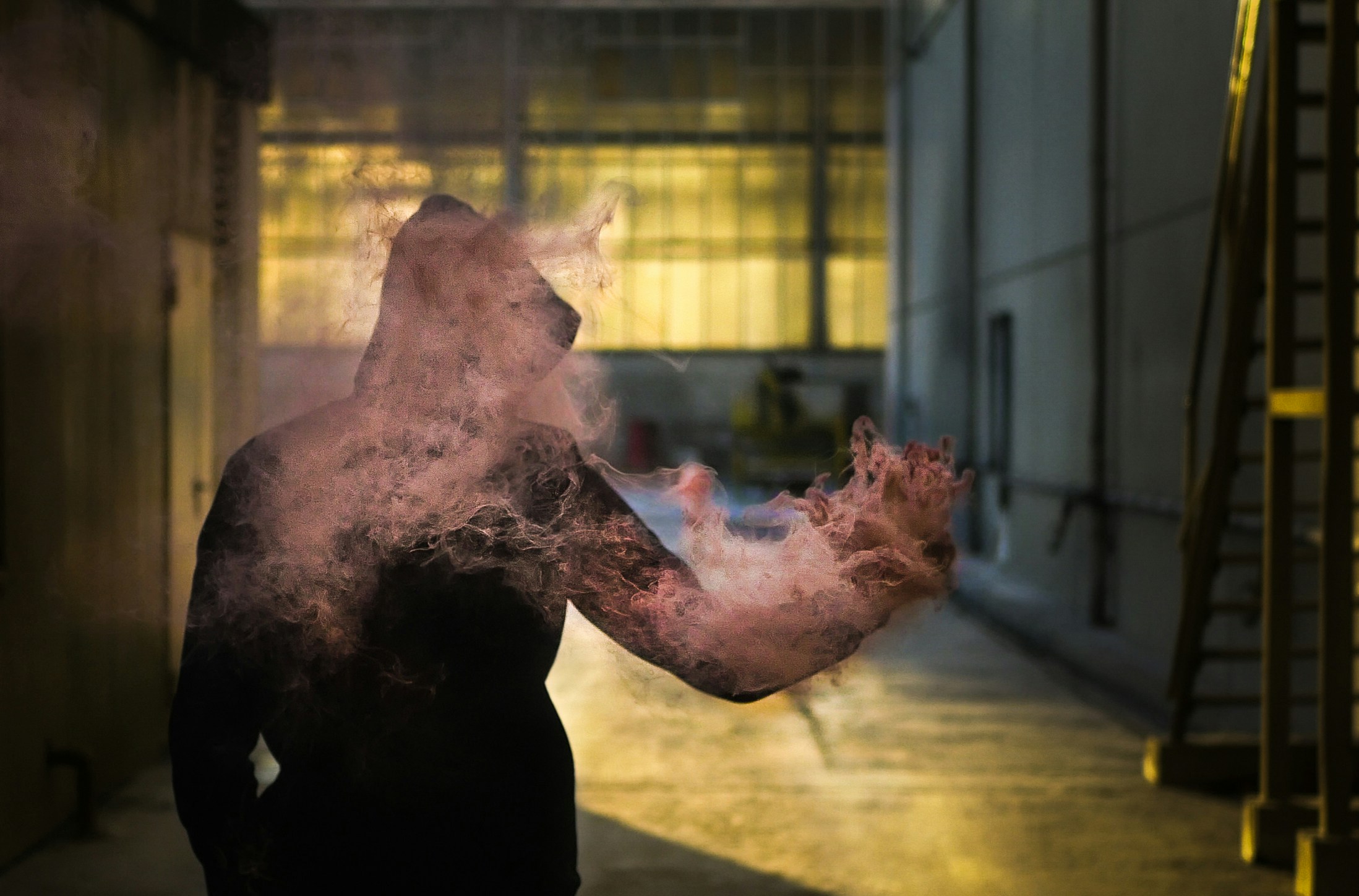 A lone silhouette in an industrial alley releases a cloud of smoke. Warm backlight from the far end silhouettes the figure and highlights the drifting particles.