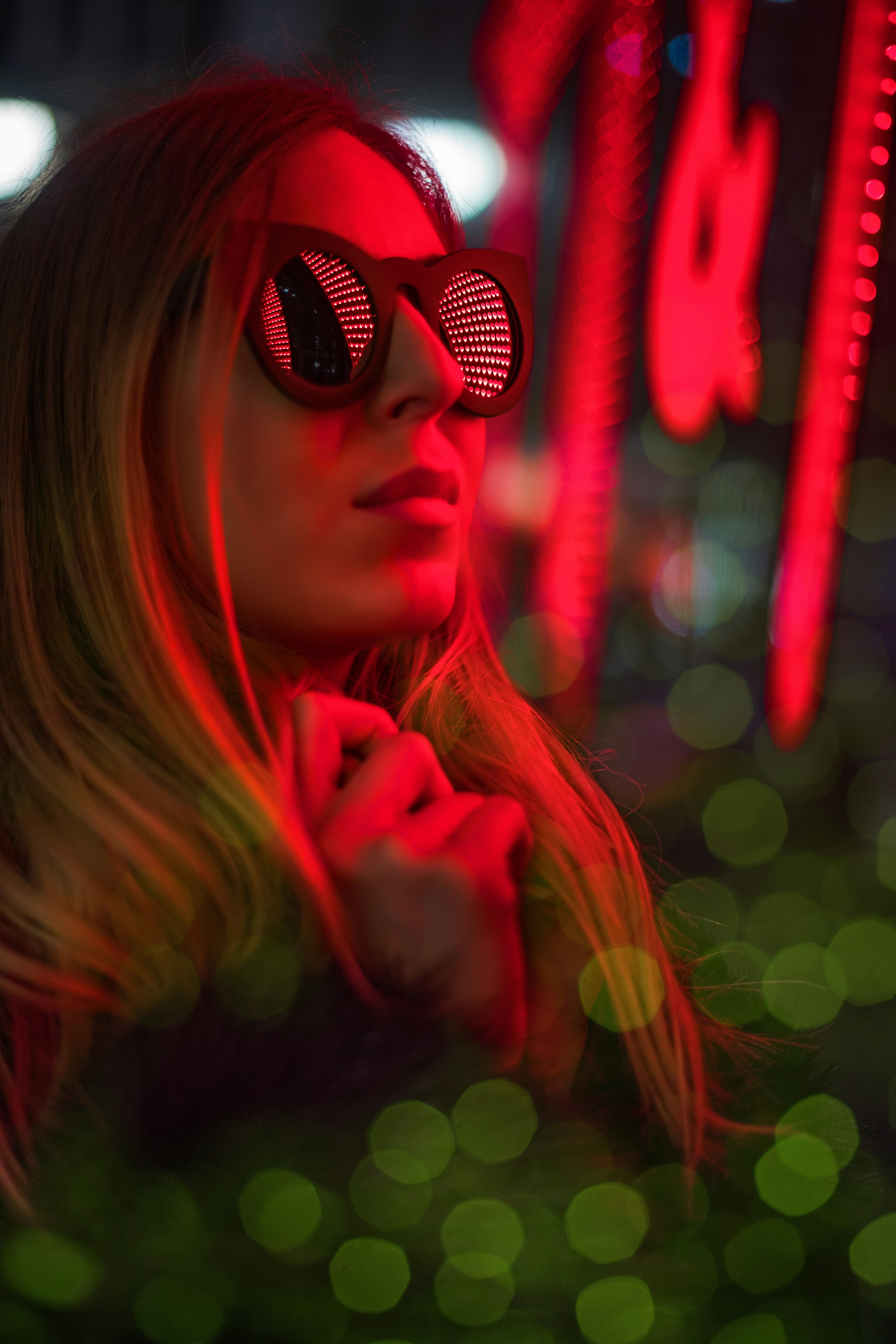 A woman with reflective sunglasses gazes thoughtfully amidst a backdrop of vibrant neon lights, creating a striking contrast of colors.