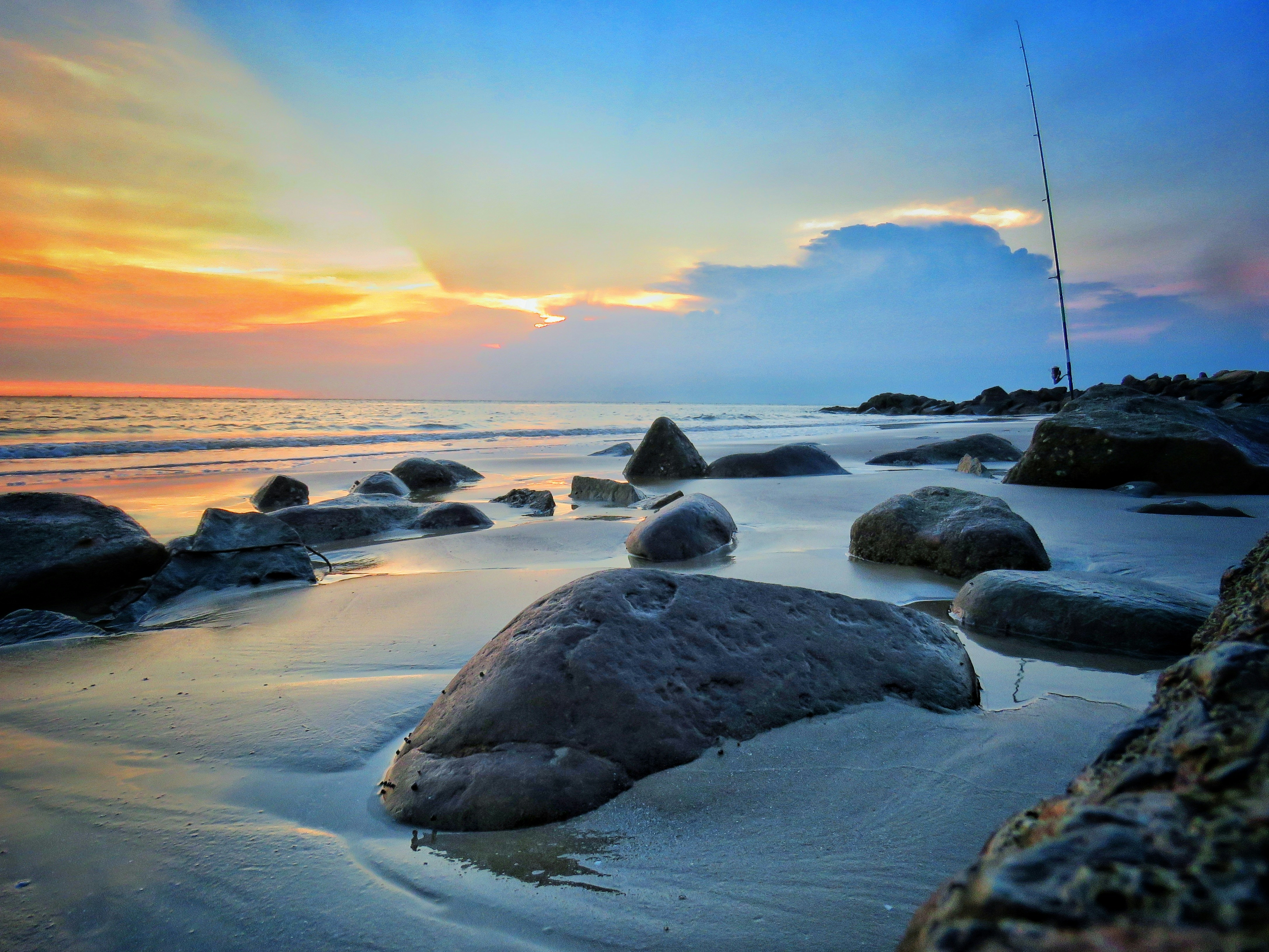 Sunset over a rocky beach with a fishing rod silhouetted against the sky.