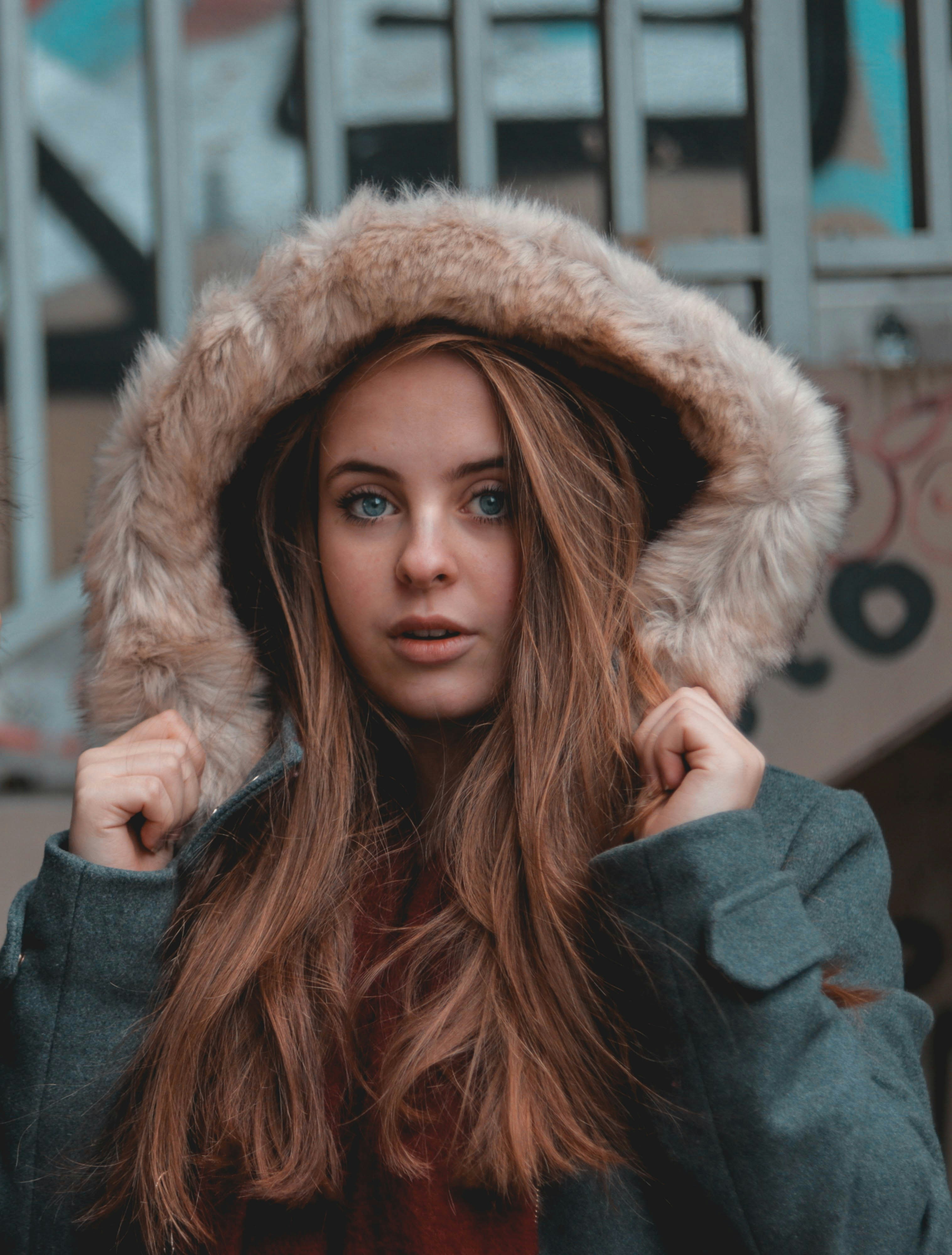 Young woman with long hair and striking blue eyes, wearing a fur-lined hooded coat, gazes thoughtfully at the camera against a colorful urban backdrop.