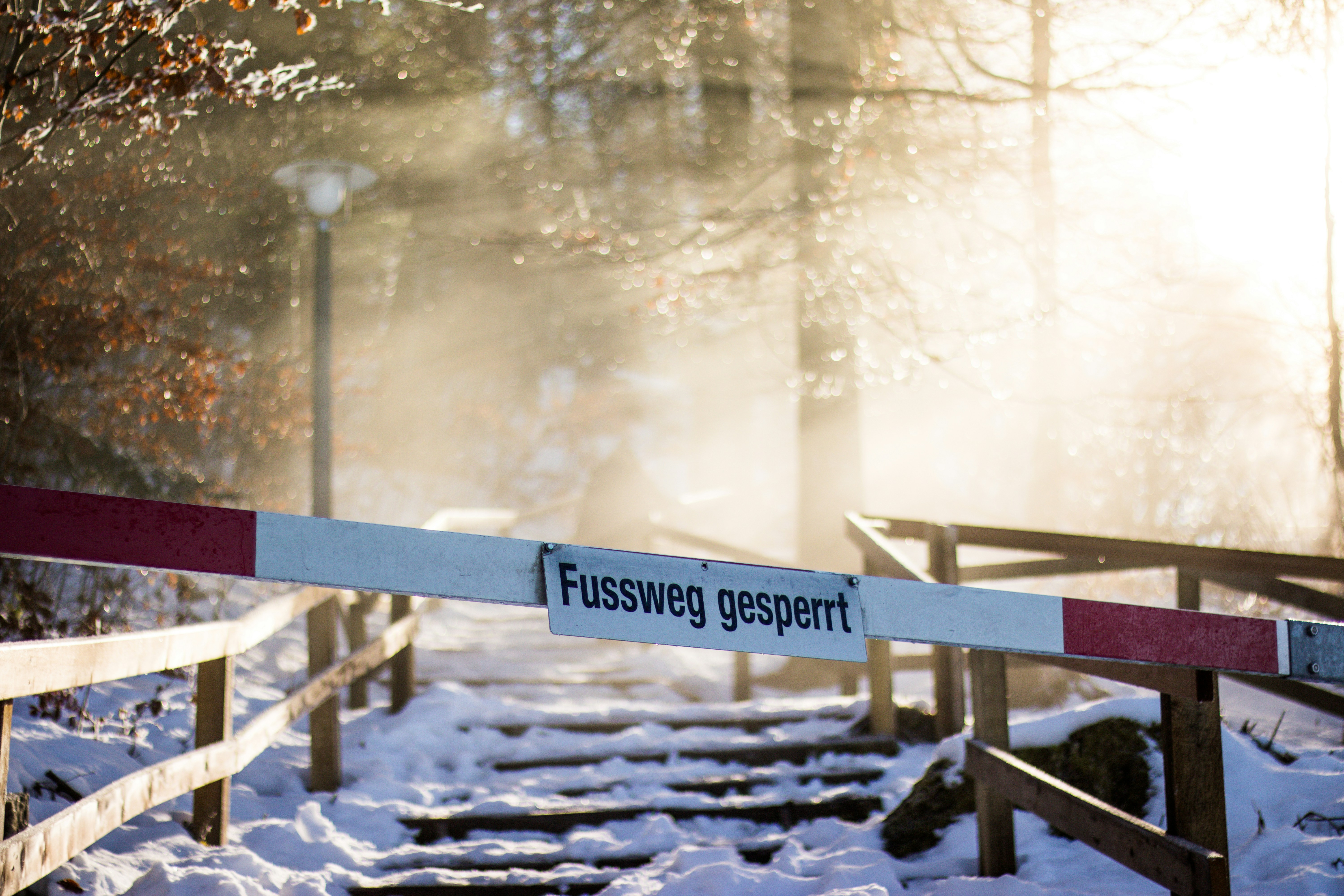 Snow-covered steps leading to a wooden barrier with the text 'Fussweg gesperrt,' indicating a closed pathway in a misty winter landscape.