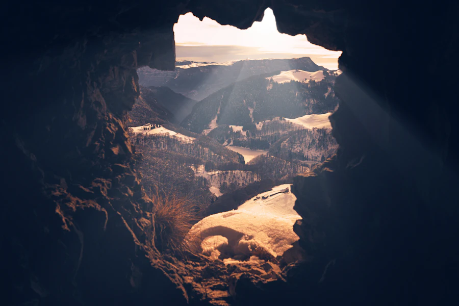 Sunlit mountain valley seen through a dark cave mouth, with snow patches and layered ridges in the distance. Warm light spills across the rocks and cave floor.