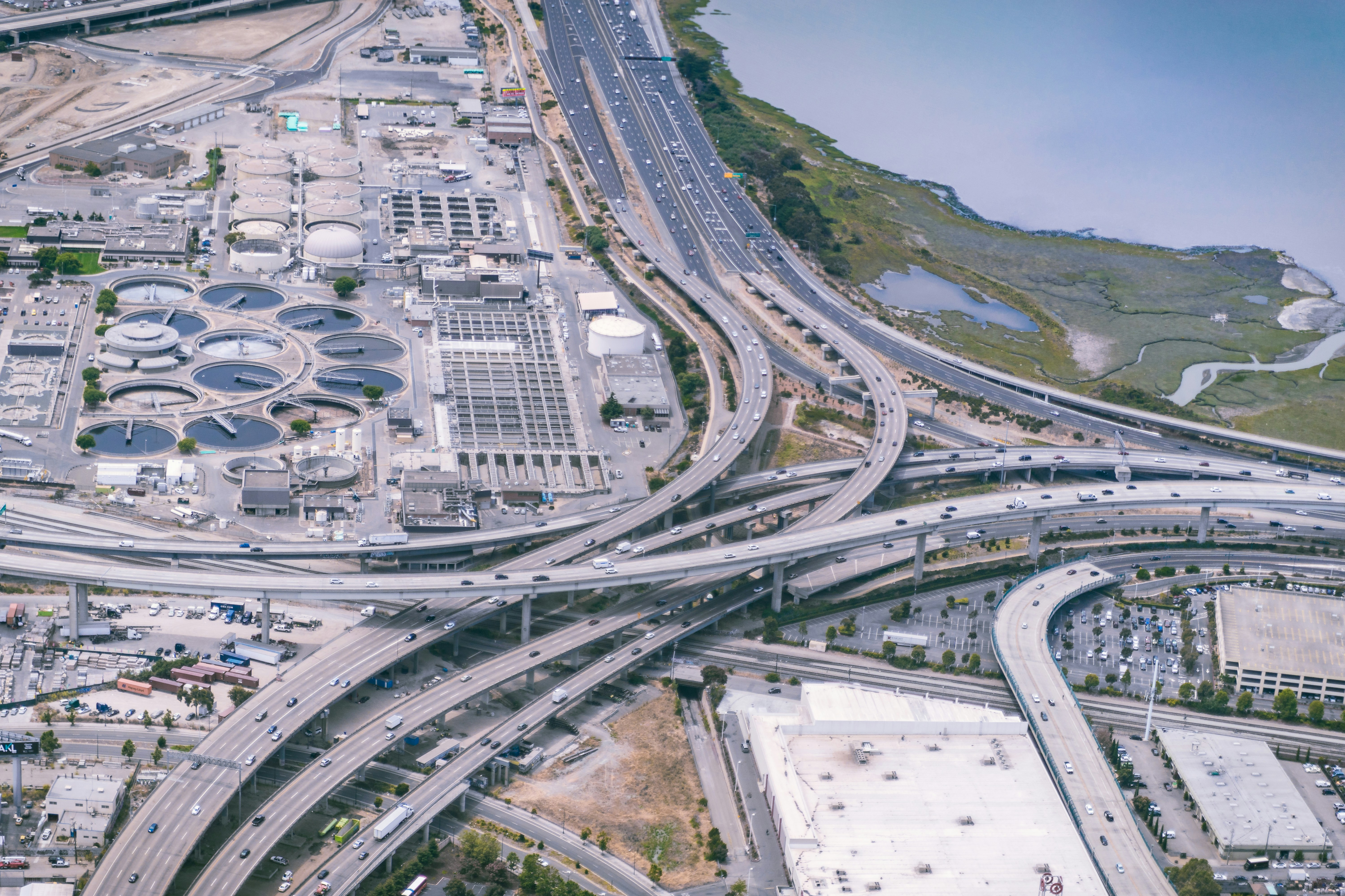 Aerial view of an intricate highway interchange with surrounding industrial structures and a nearby water body.