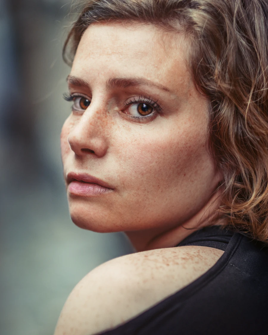 Close-up portrait of a woman looking over her shoulder, with soft natural light highlighting her eyes and freckled skin. Blurred background keeps attention on her face and expression.