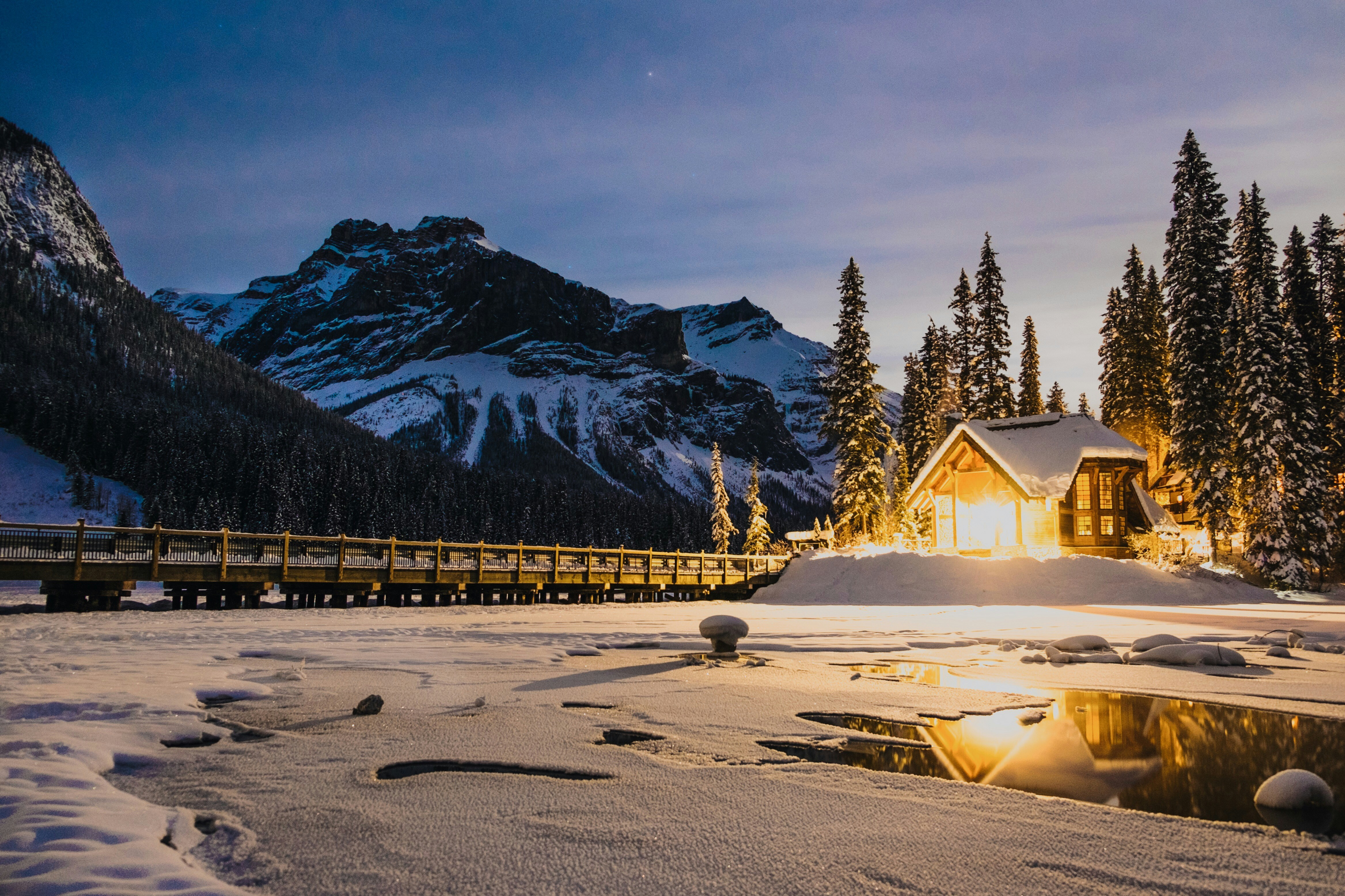 Cozy cabin illuminated at dusk against a snow-covered mountain backdrop.
