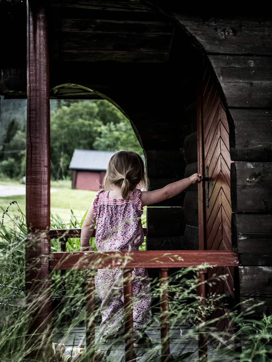 A girl in a floral dress stands with her back to the camera inside a weathered wooden playhouse, her hand on the door as greenery and a distant red shed blur in the background.