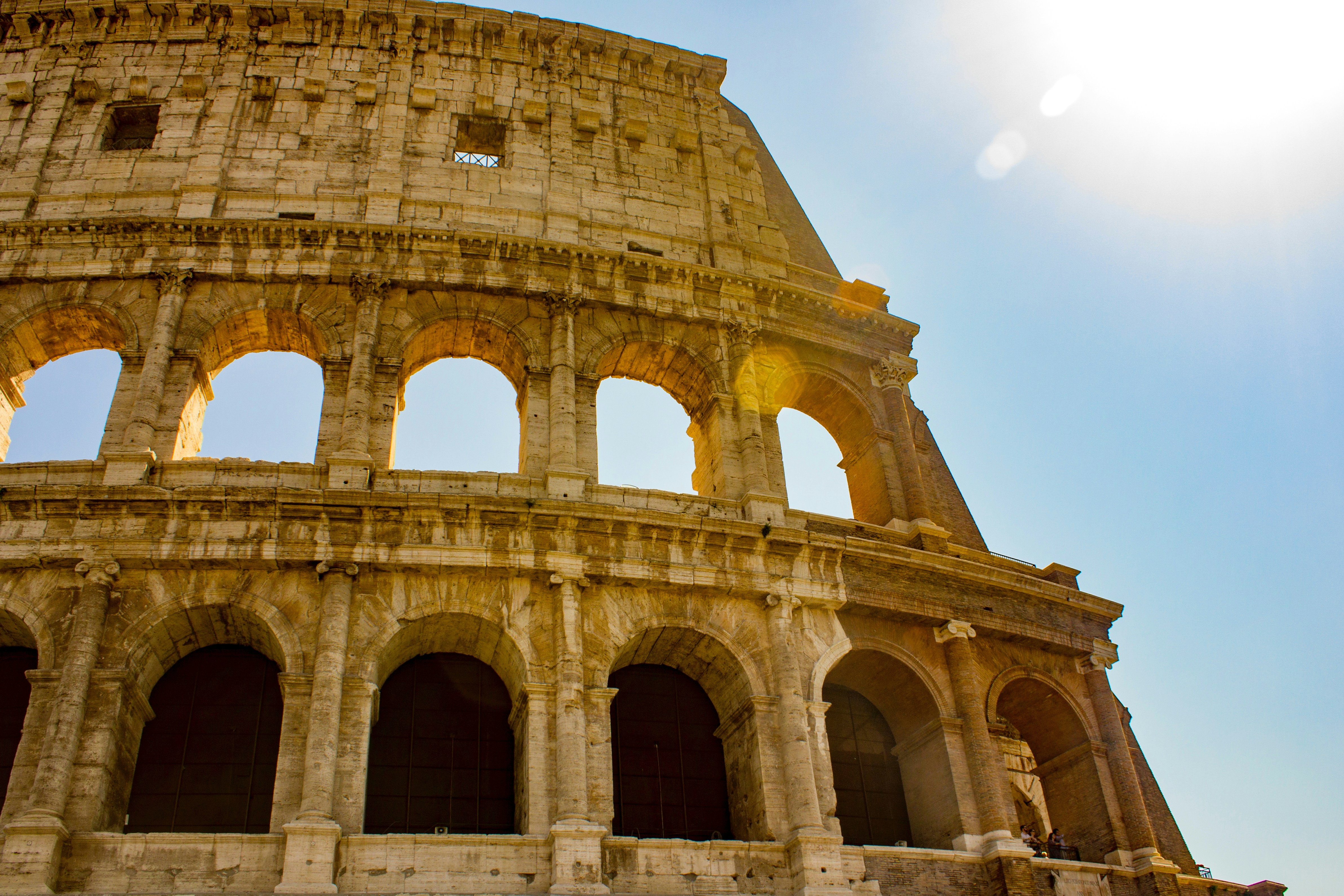 The Colosseum's towering arches bathed in warm sunlight with a clear blue sky.