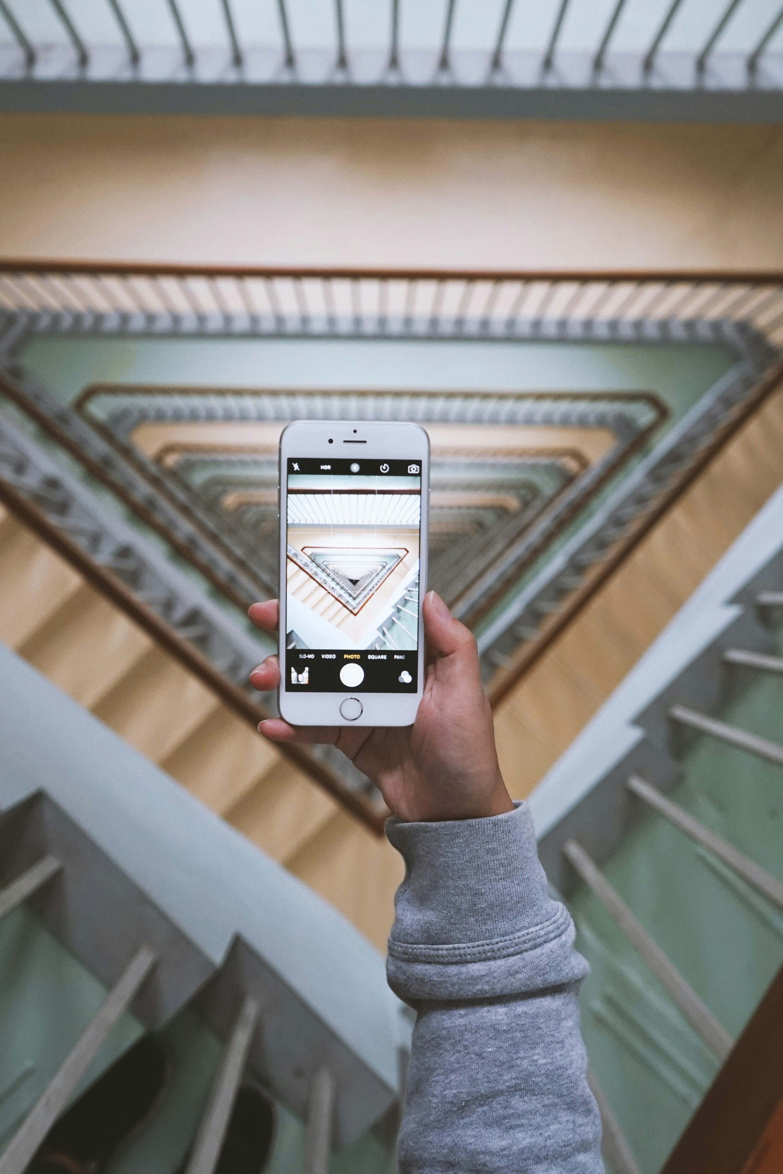 A hand holds a smartphone displaying a view of a spiraling staircase, creating a visual echo of geometric patterns. The composition emphasizes depth and symmetry.