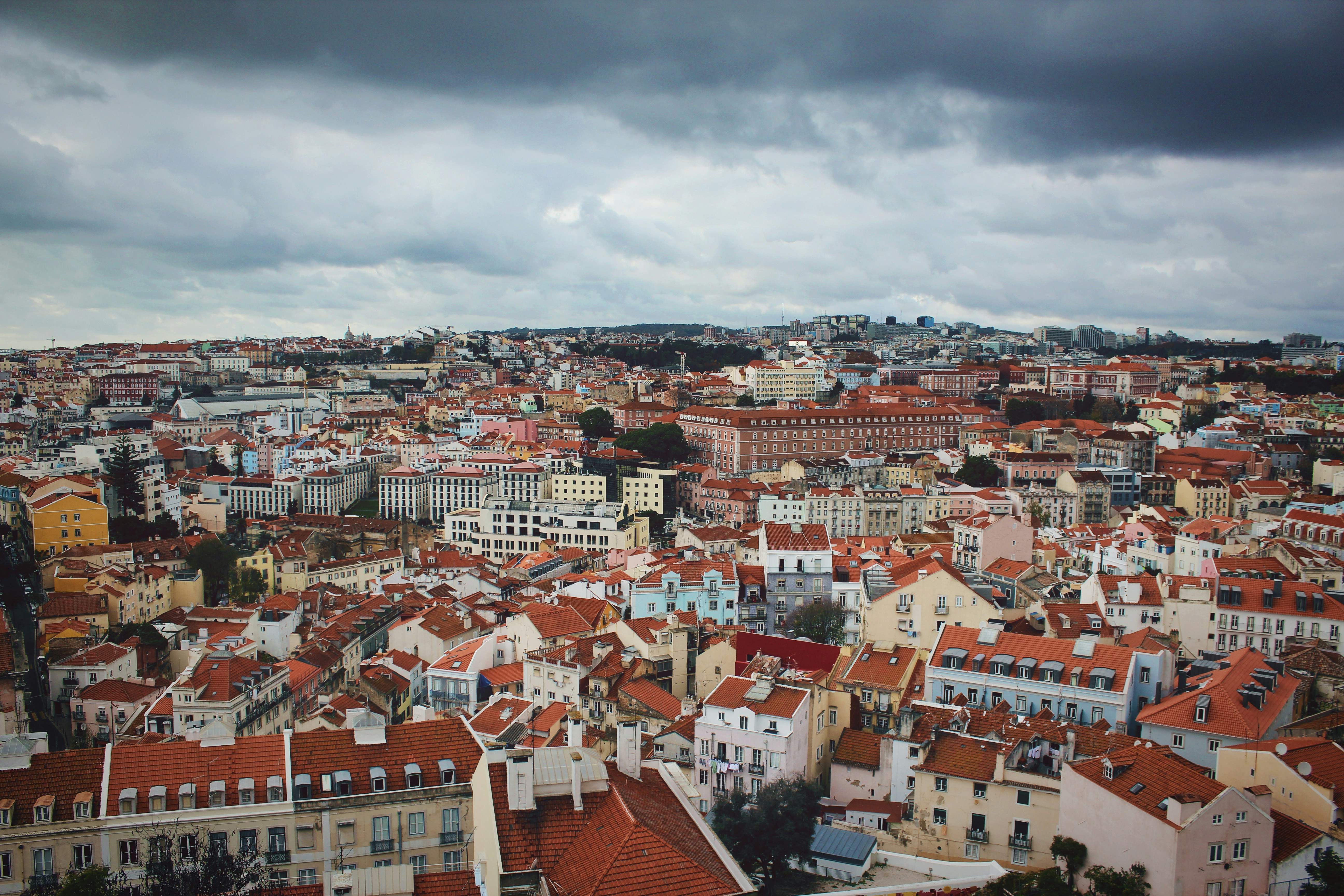 A panoramic view of Lisbon showcasing a dense cluster of buildings with distinctive terracotta roofs under a moody sky.