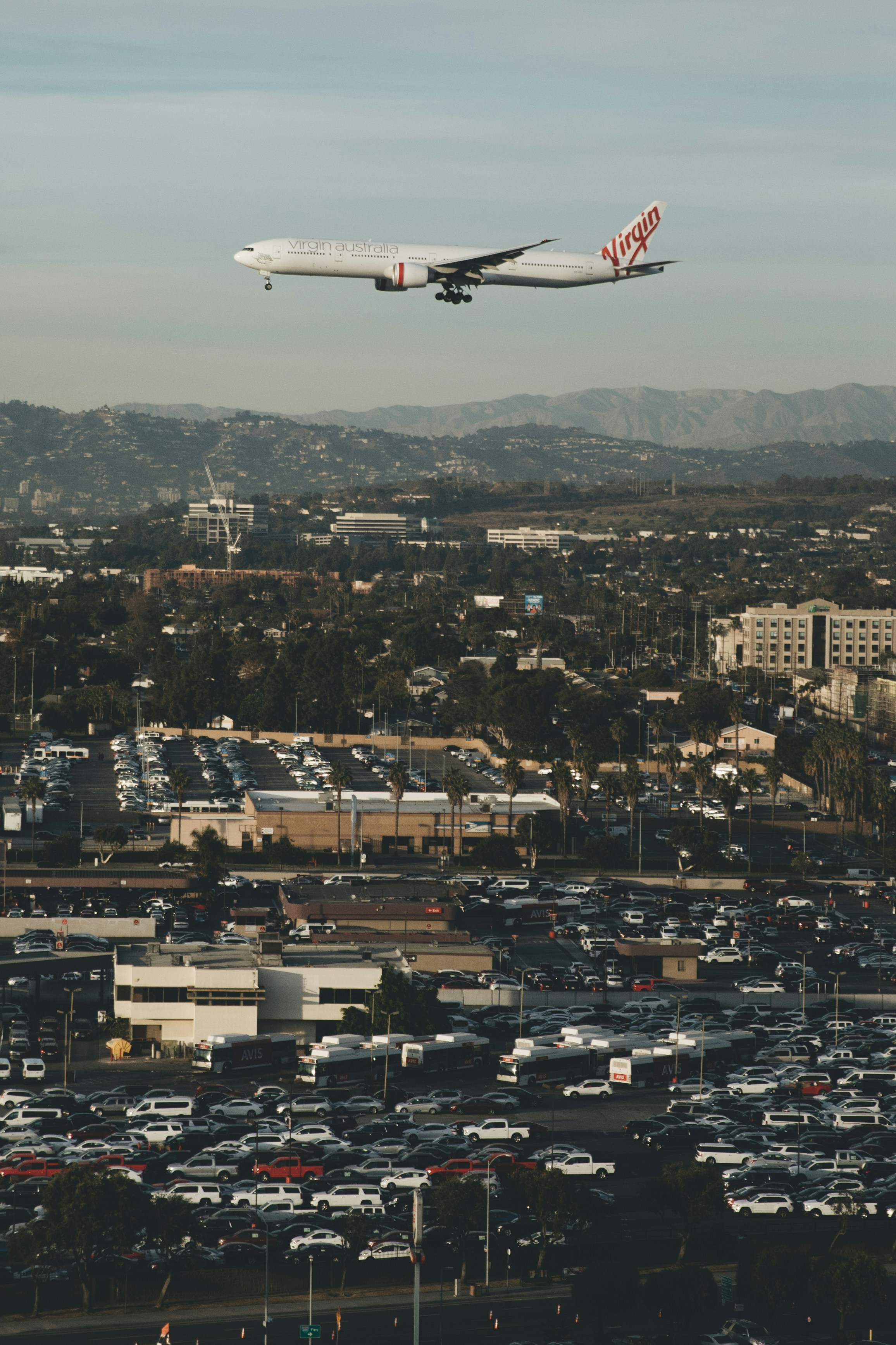 A Virgin Atlantic aircraft descends gracefully towards the airport, framed by a sprawling urban parking lot and distant hills. 
