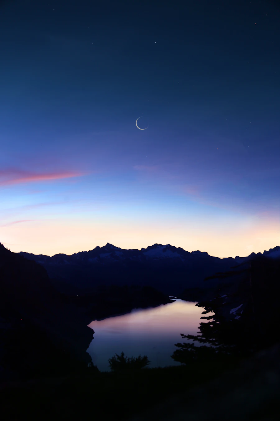 Thin crescent moon above a mountain range at dawn, with a pastel sky fading from deep blue to warm peach. A calm lake below reflects the fading light.